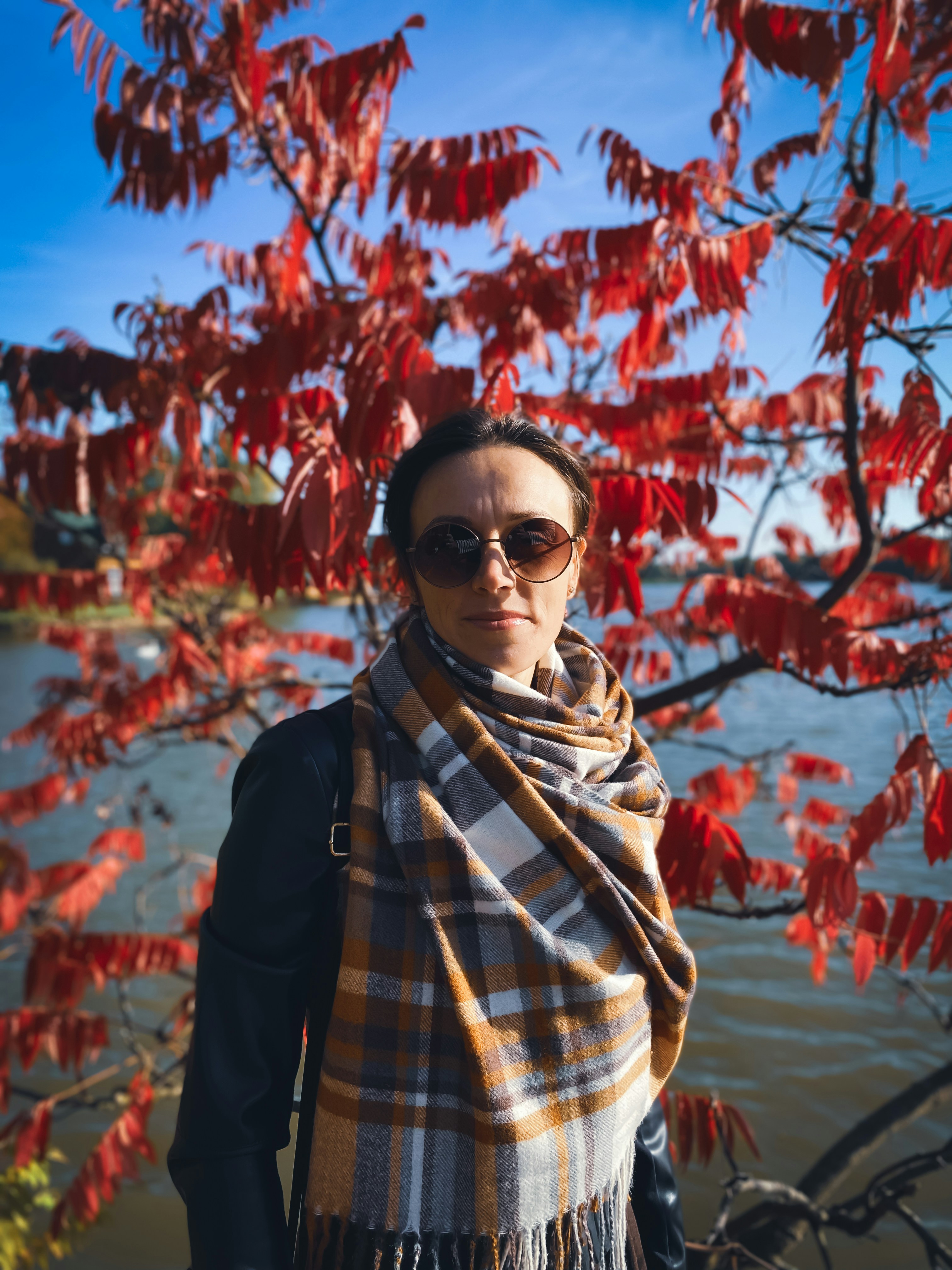 Portrait of a person wearing sunglasses and a plaid scarf, standing in front of red autumn leaves by a body of water.