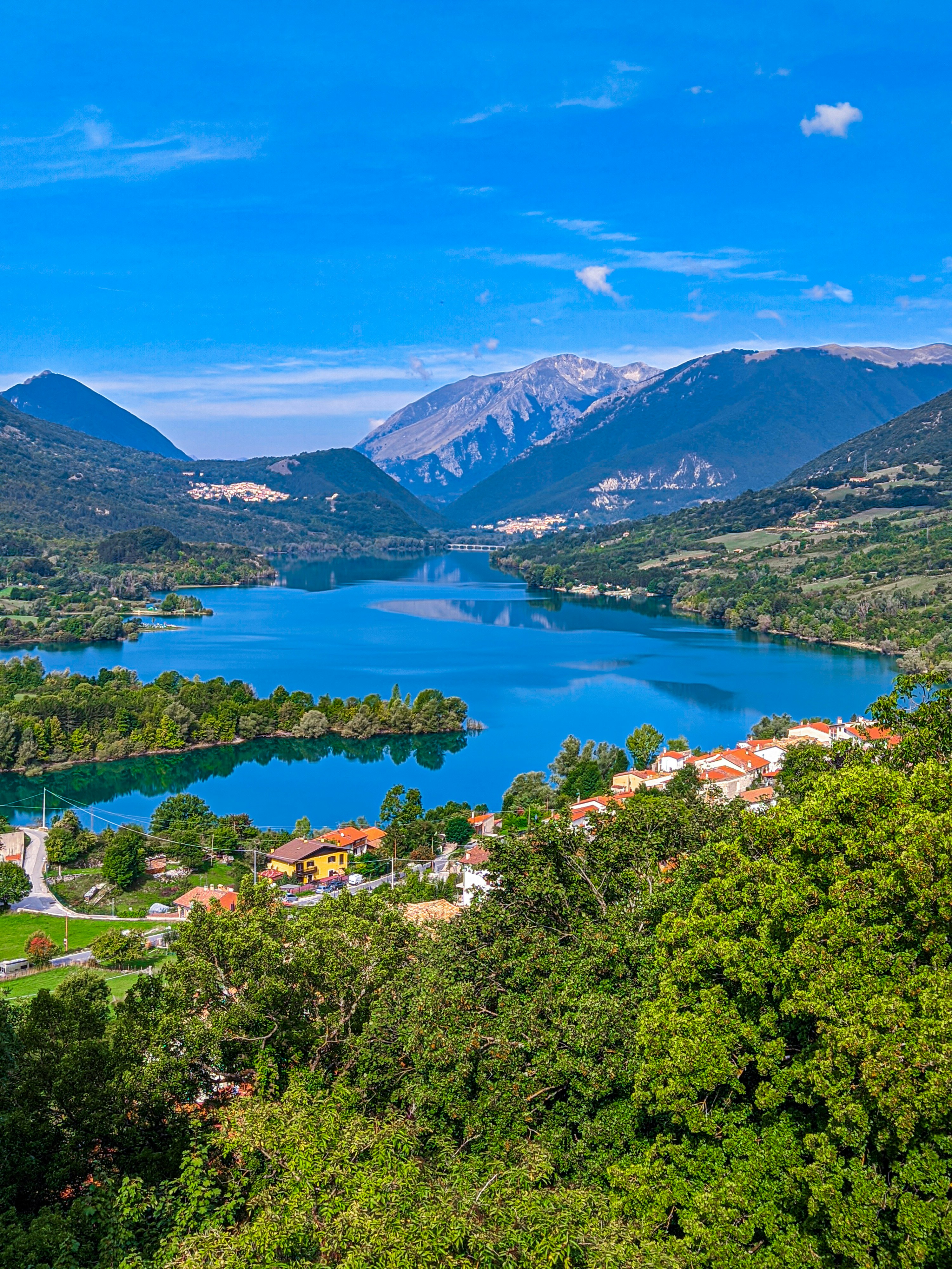 Landscape photograph of a turquoise lake winding through a valley, framed by green hills and a cluster of red-roofed houses, with towering mountains in the distance.