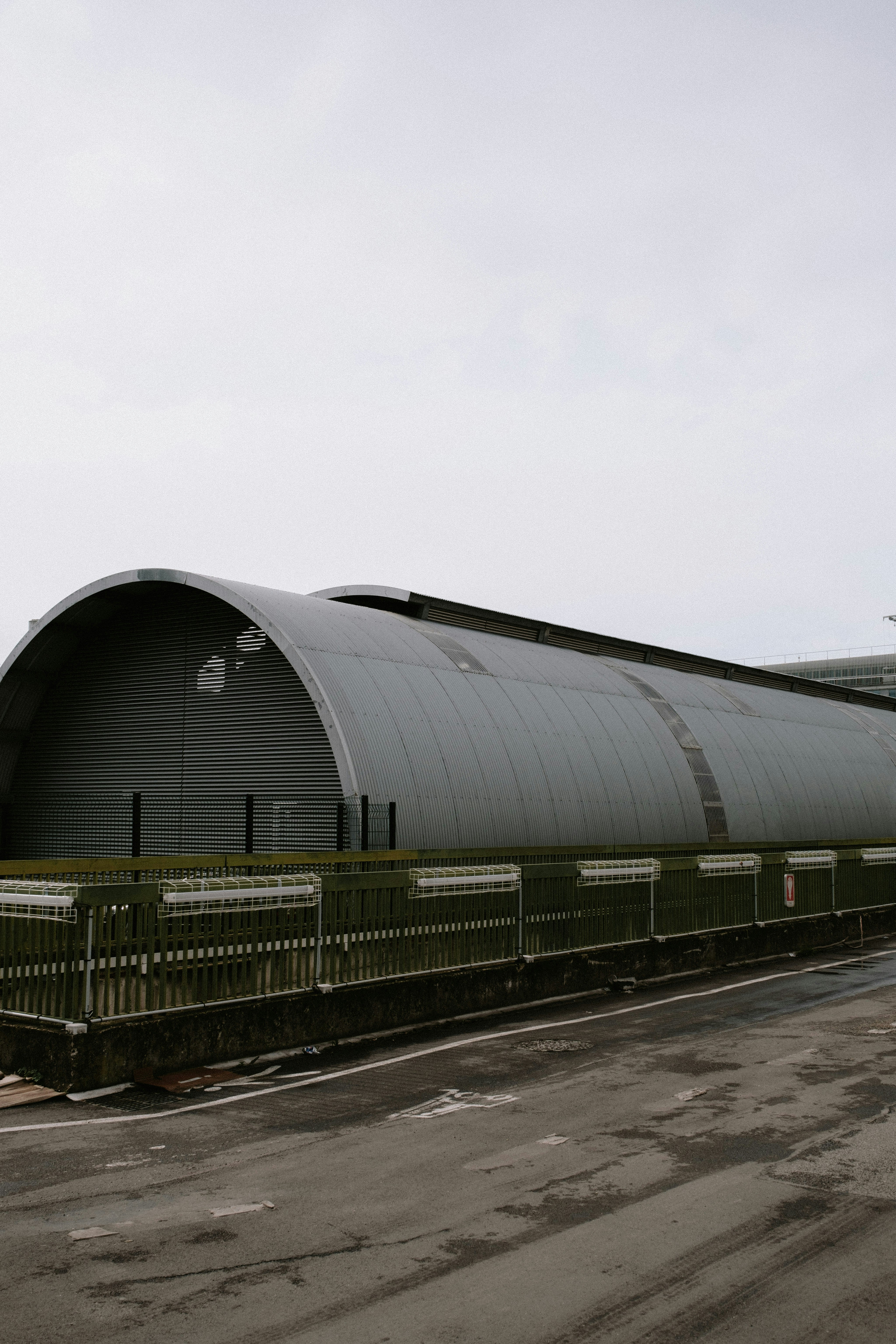 Sleek, curved building structure with metallic facade and horizontal slats, set against a muted sky. The surrounding area features a green fence and asphalt pavement.