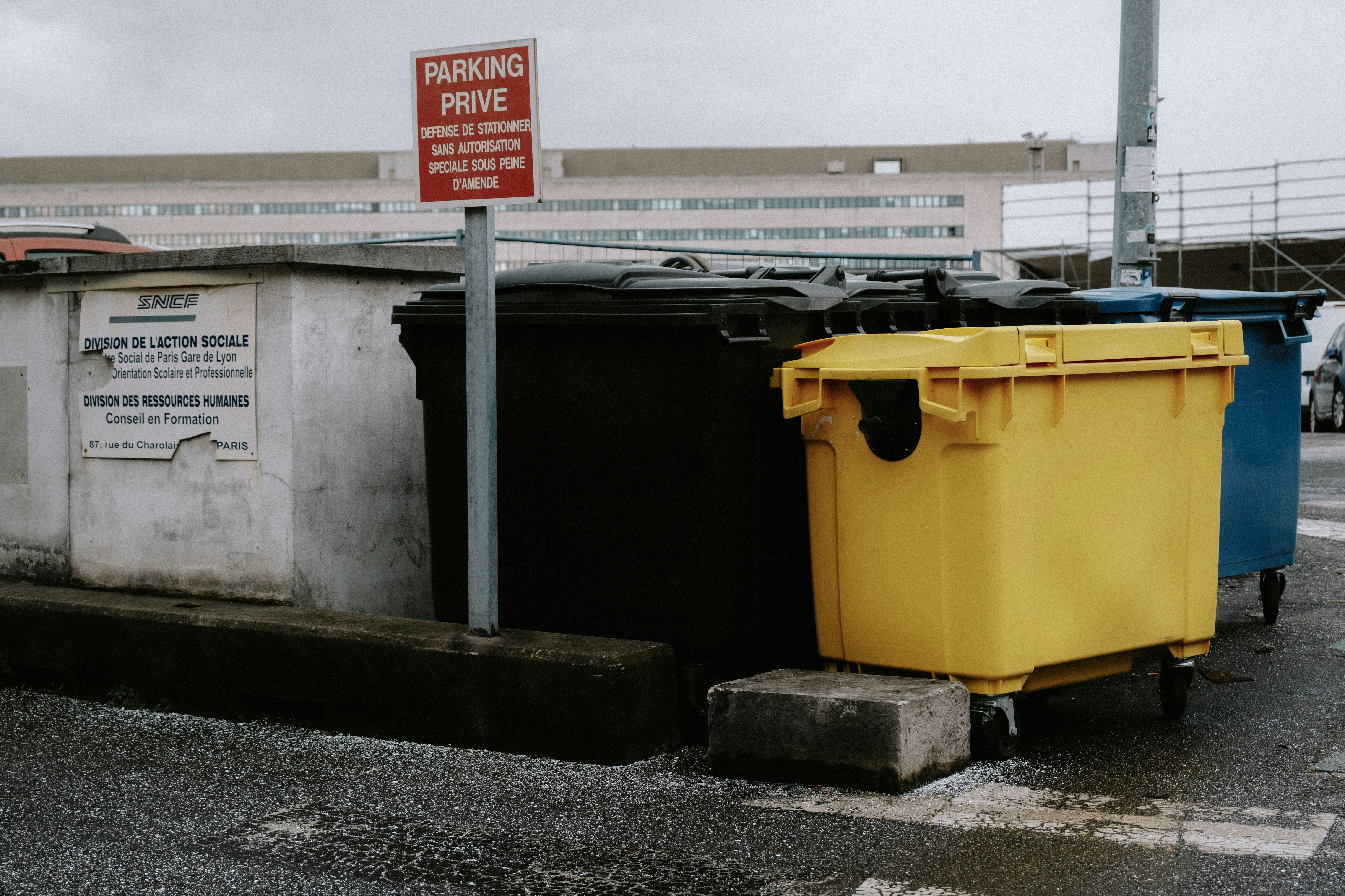 A collection of waste containers in a parking area, showcasing a vibrant yellow bin among darker counterparts. The scene captures the intersection of urban life and environmental awareness.