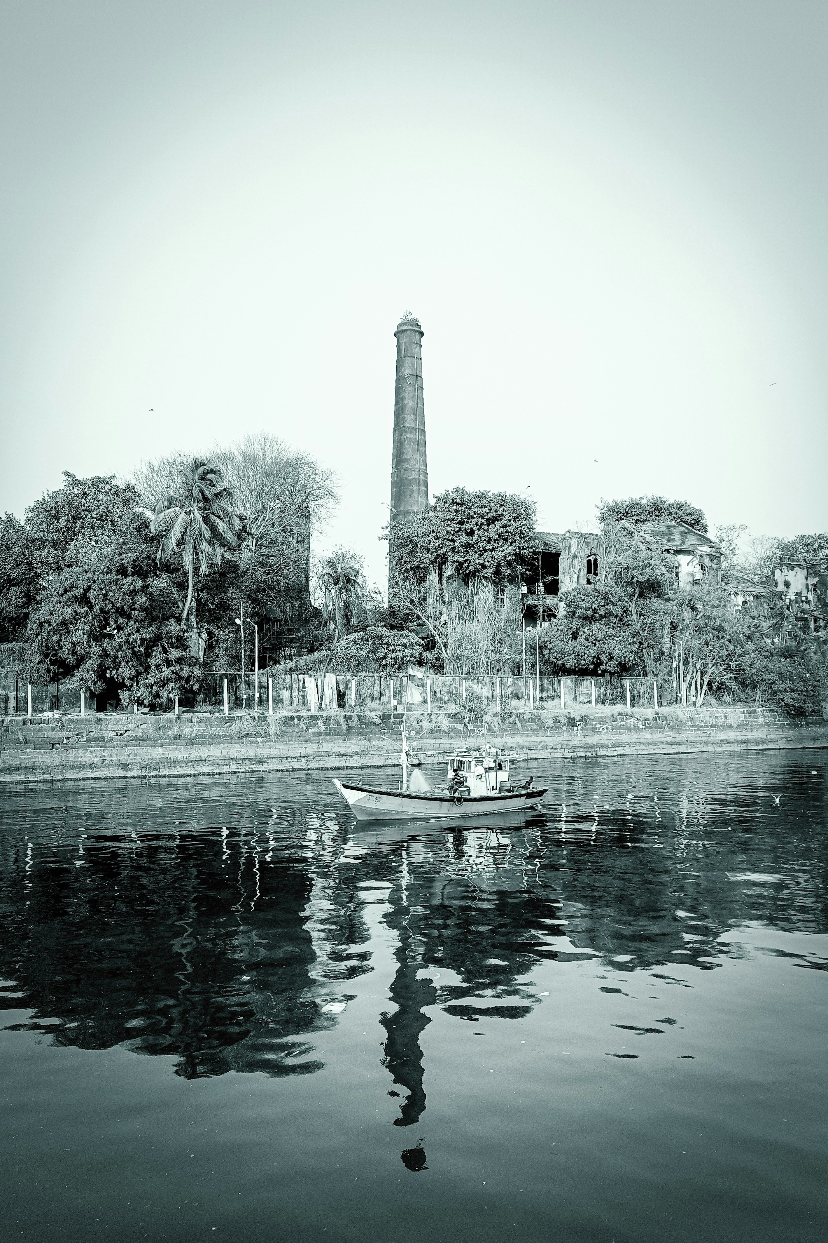 Black-and-white photograph of a riverfront scene with a small boat gliding on calm water and a tall chimney framed by trees along the shore.