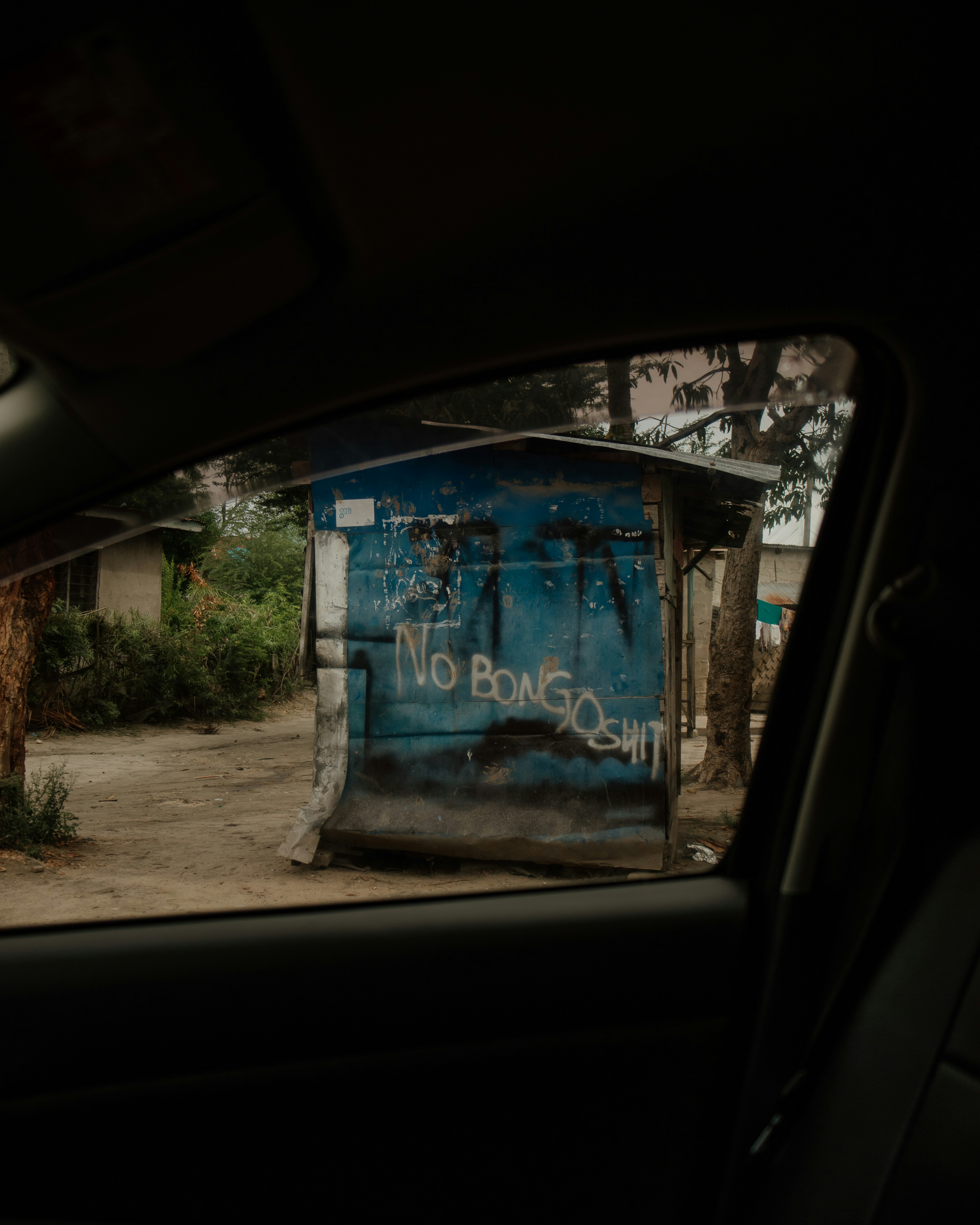 Graffiti-covered shed in an urban setting, partially framed by a car window. The message 'No Bongo Shit' stands out against the blue backdrop.
