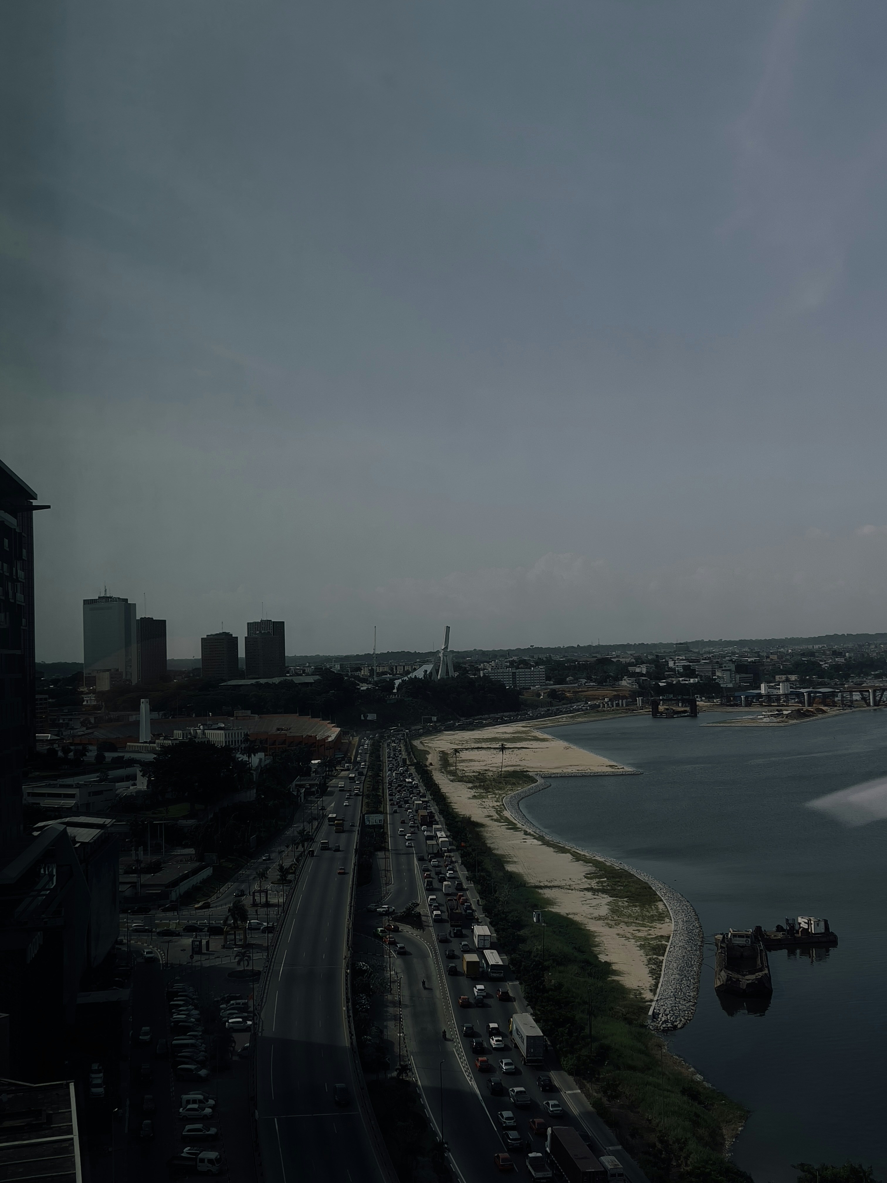 Aerial view of a bustling cityscape alongside a serene waterfront, showcasing a busy road and sandy beach. The contrast between urban life and nature is evident.