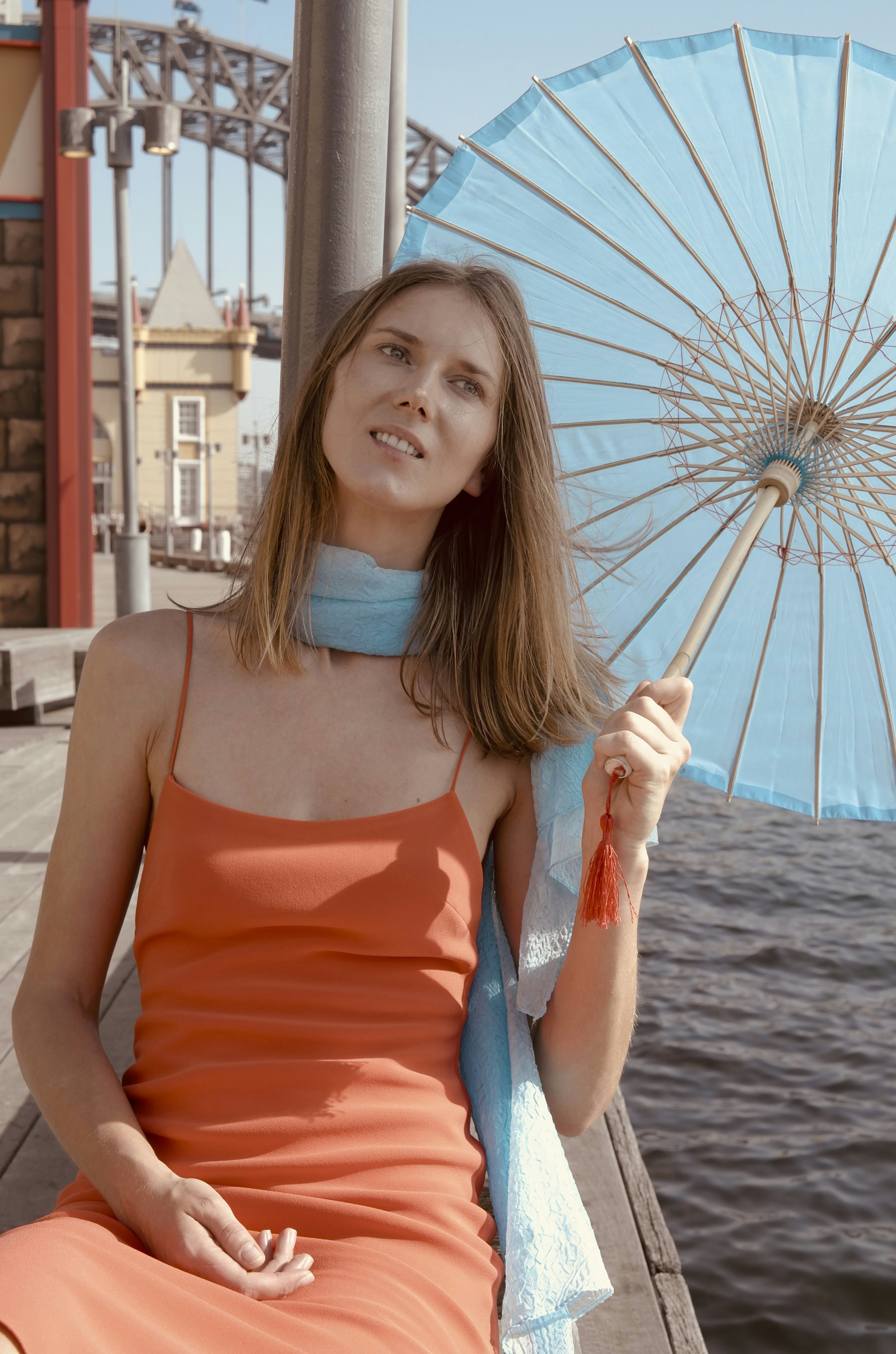 Portrait photograph of a woman in an orange dress sitting on a wooden pier, holding a blue parasol by the water.