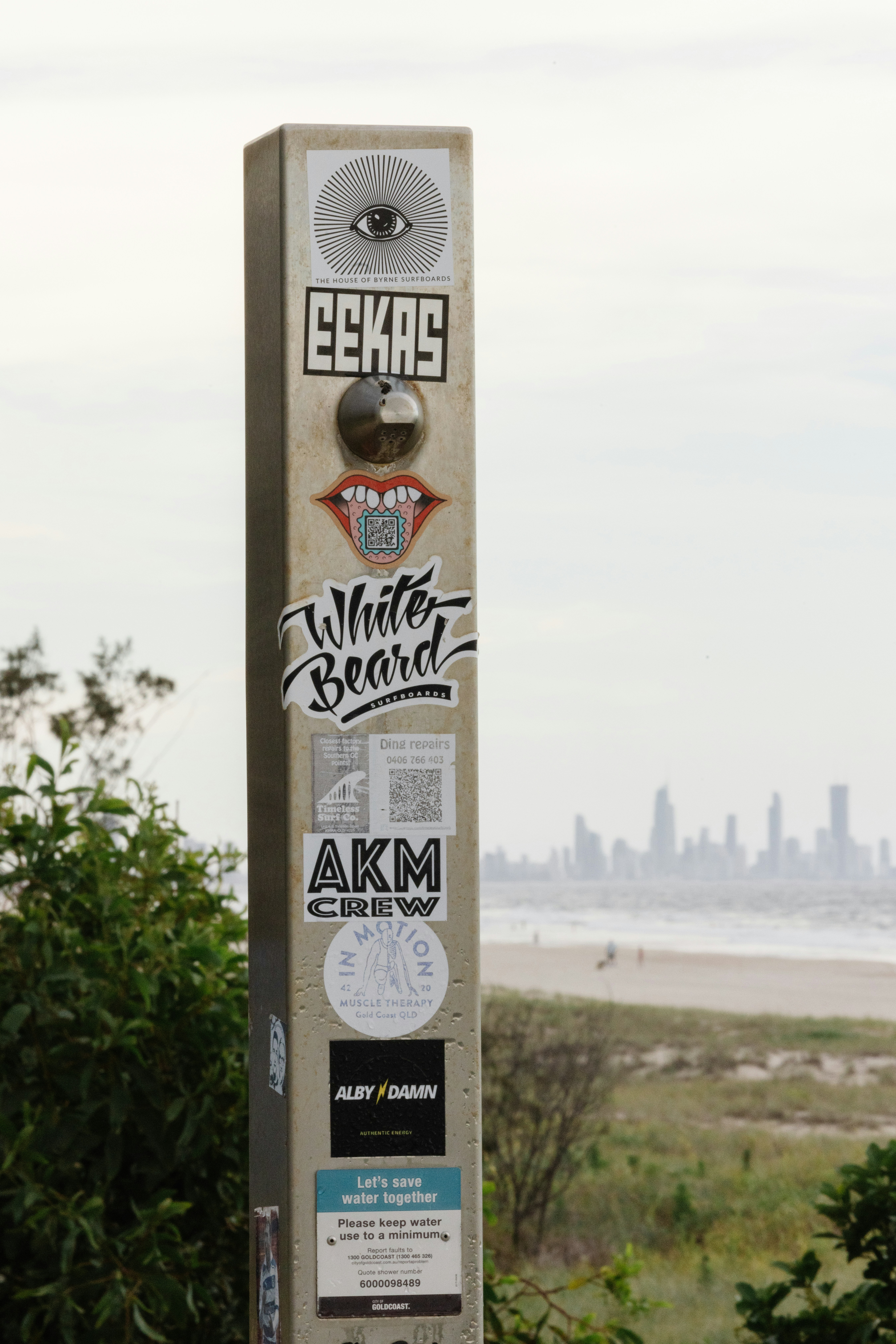 A weathered pole adorned with various stickers and graffiti stands against a backdrop of a distant city skyline along the beach. The scene captures a blend of urban art and natural beauty.