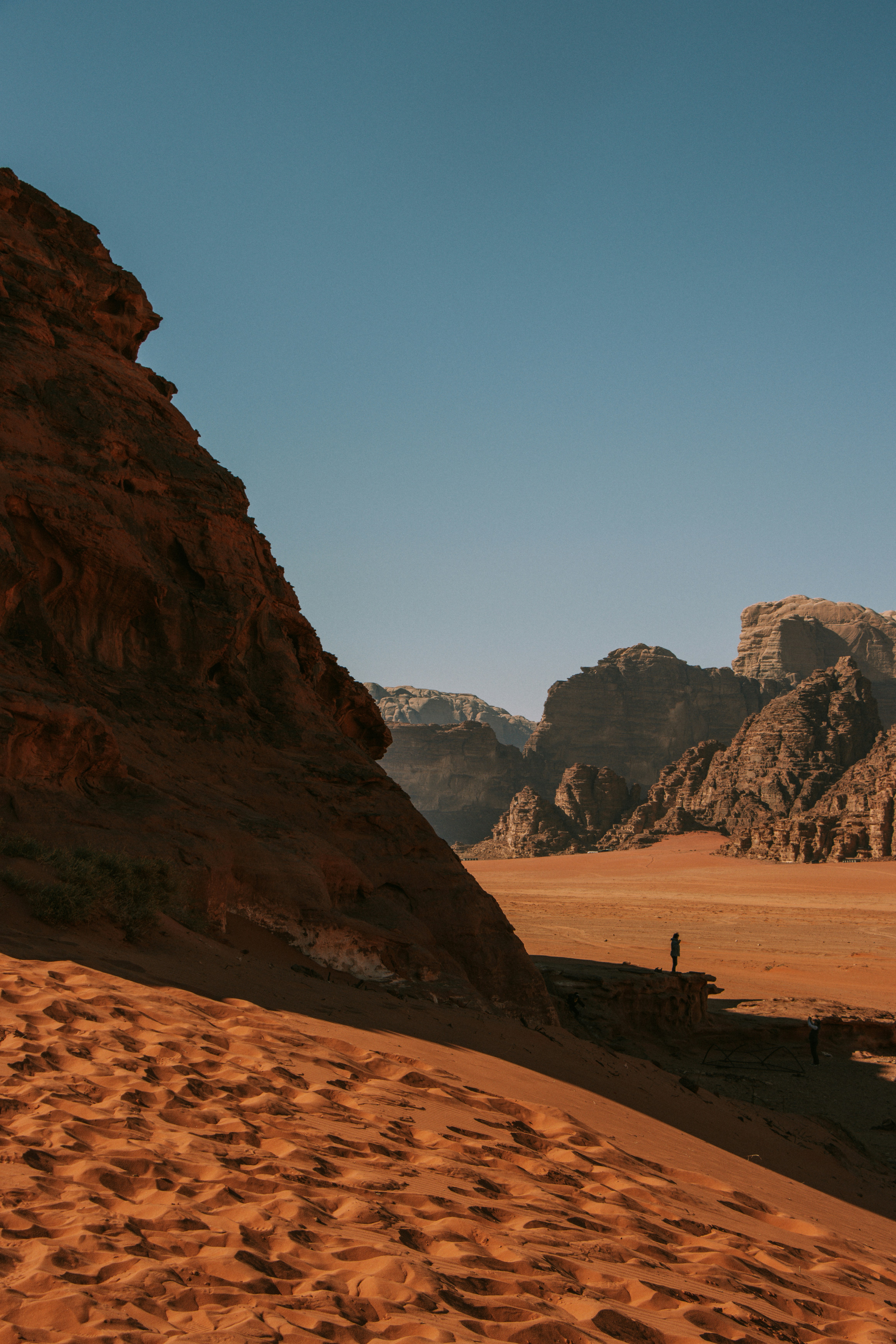 Lone figure traverses expansive desert landscape surrounded by towering rock formations under a clear blue sky.