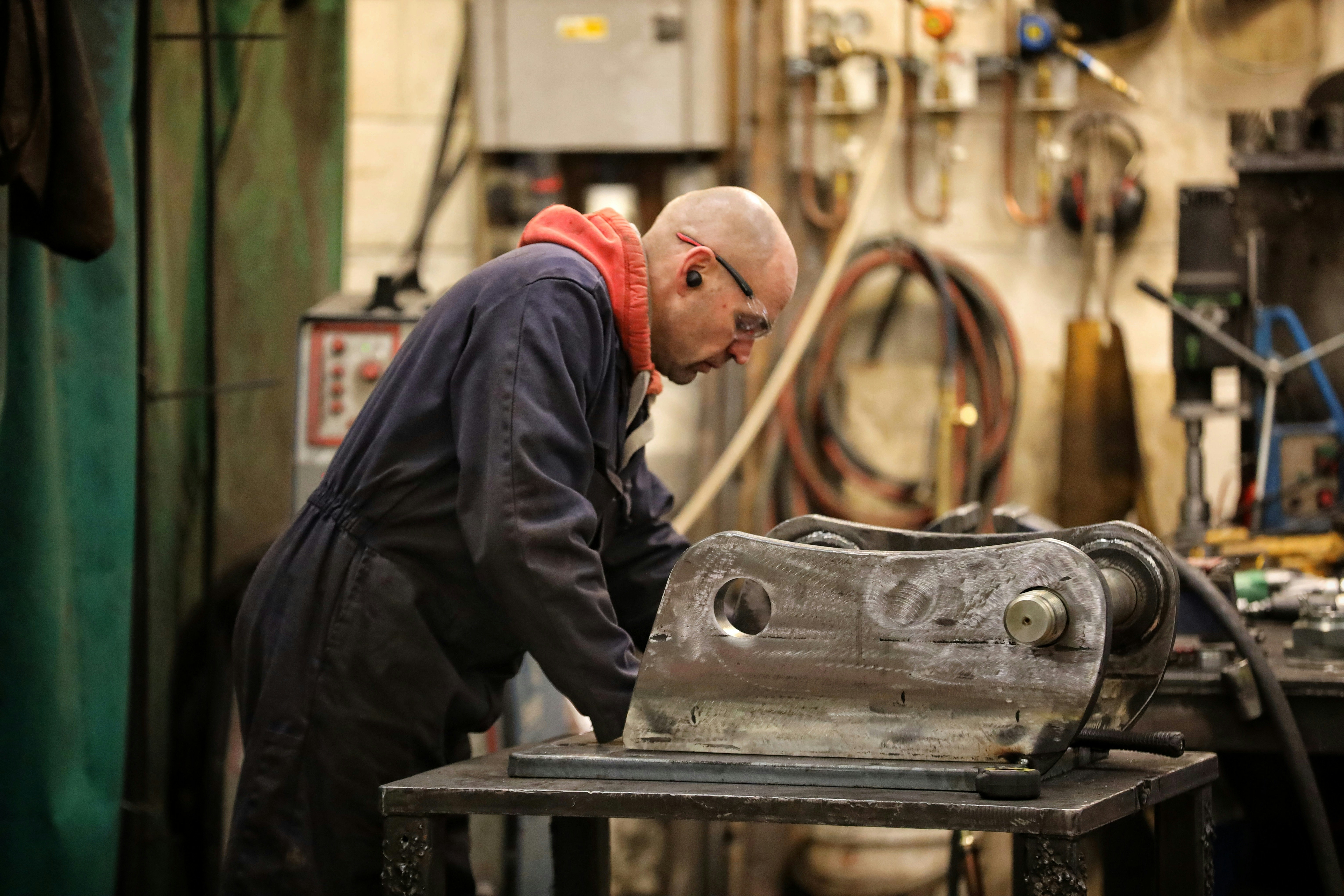A skilled worker meticulously examines a metal piece in a workshop, surrounded by tools and machinery. The focus is on the craftsmanship involved in metal fabrication.