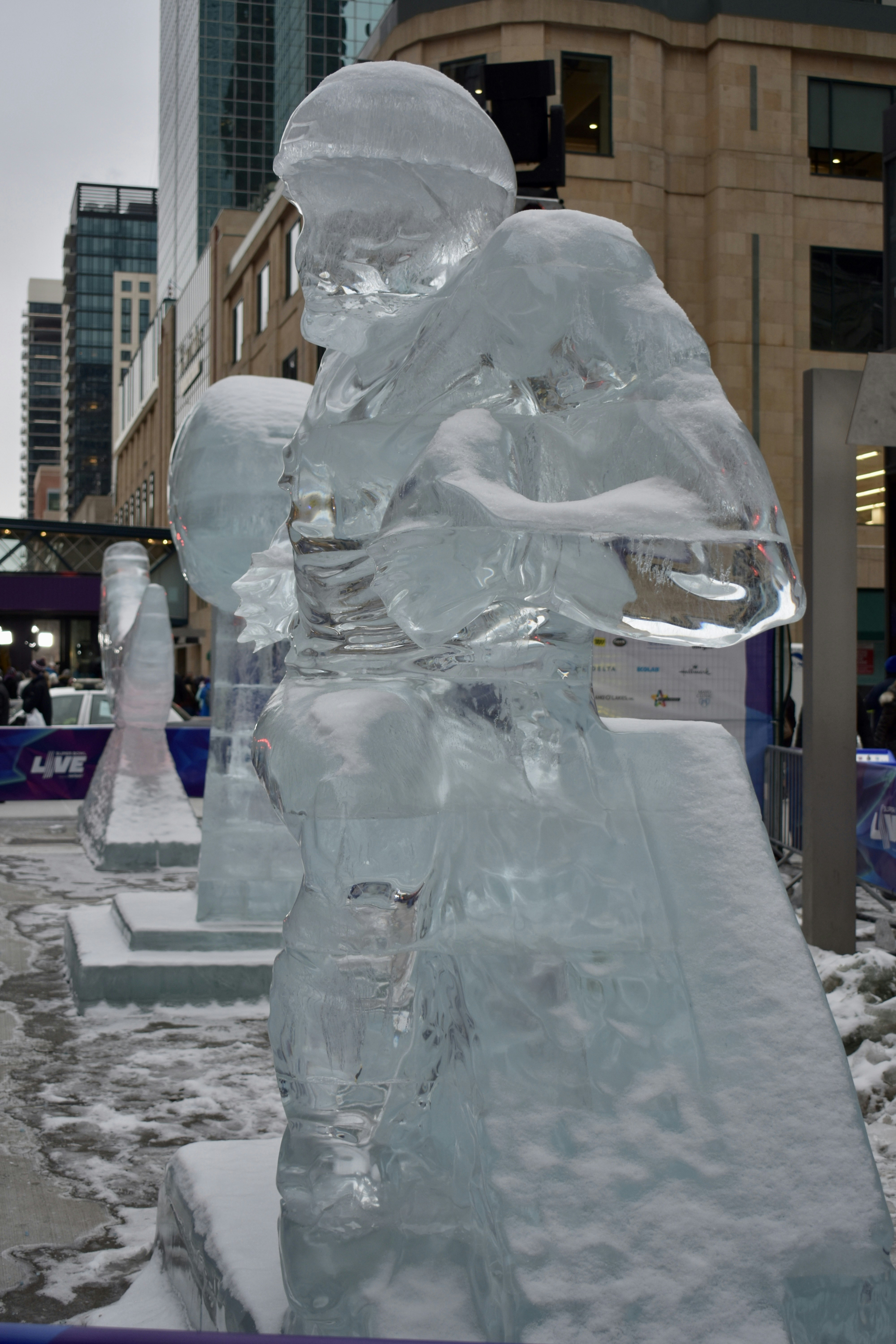 Intricately carved ice sculpture of a football player in action, surrounded by a wintery urban backdrop. Snow covers the ground, enhancing the chilly atmosphere.