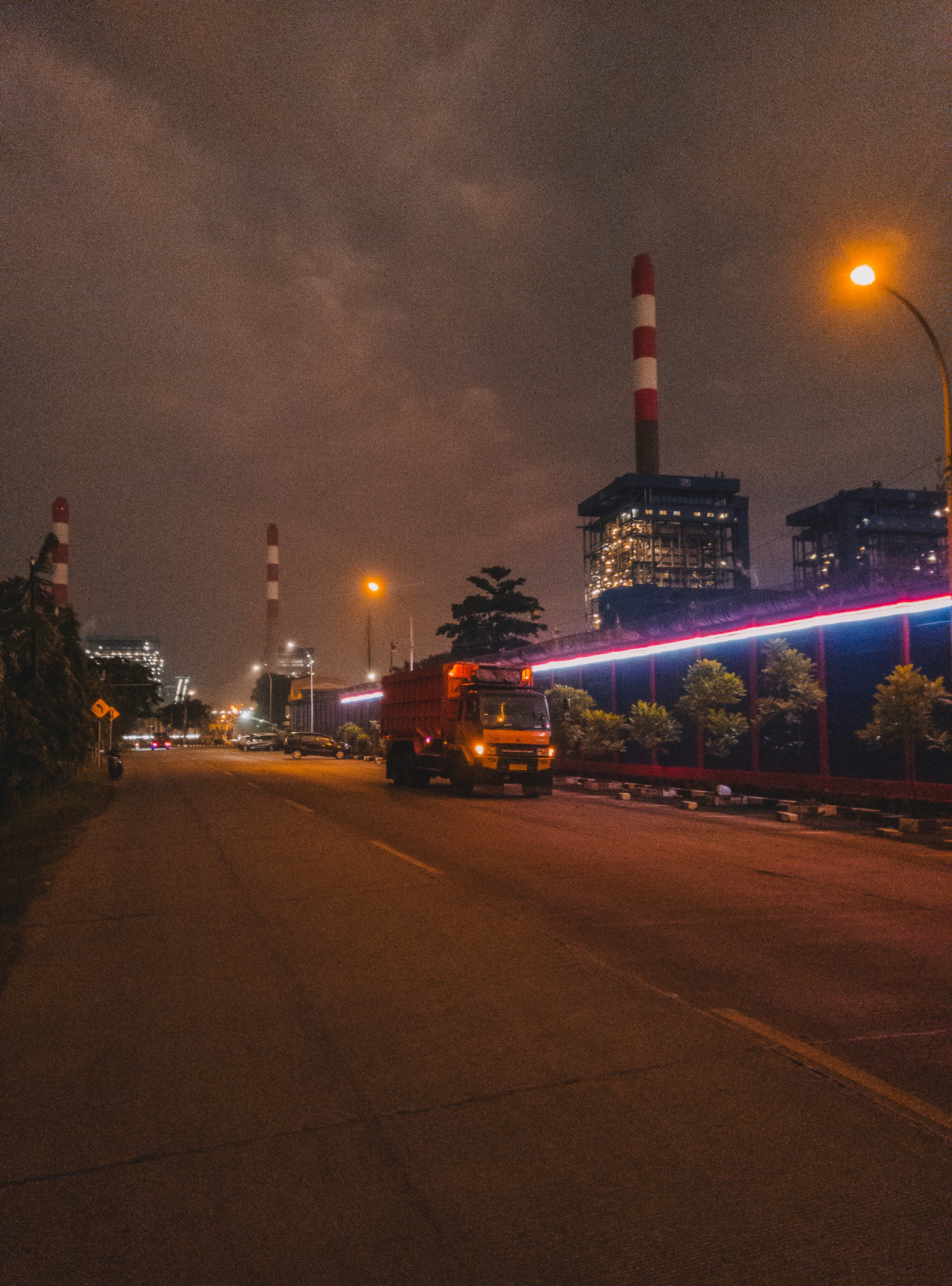 Dimly lit street scene at night featuring a truck and distant factory chimneys with red-white stripes; a neon light trail runs along a fence, highlighting an industrial area.
