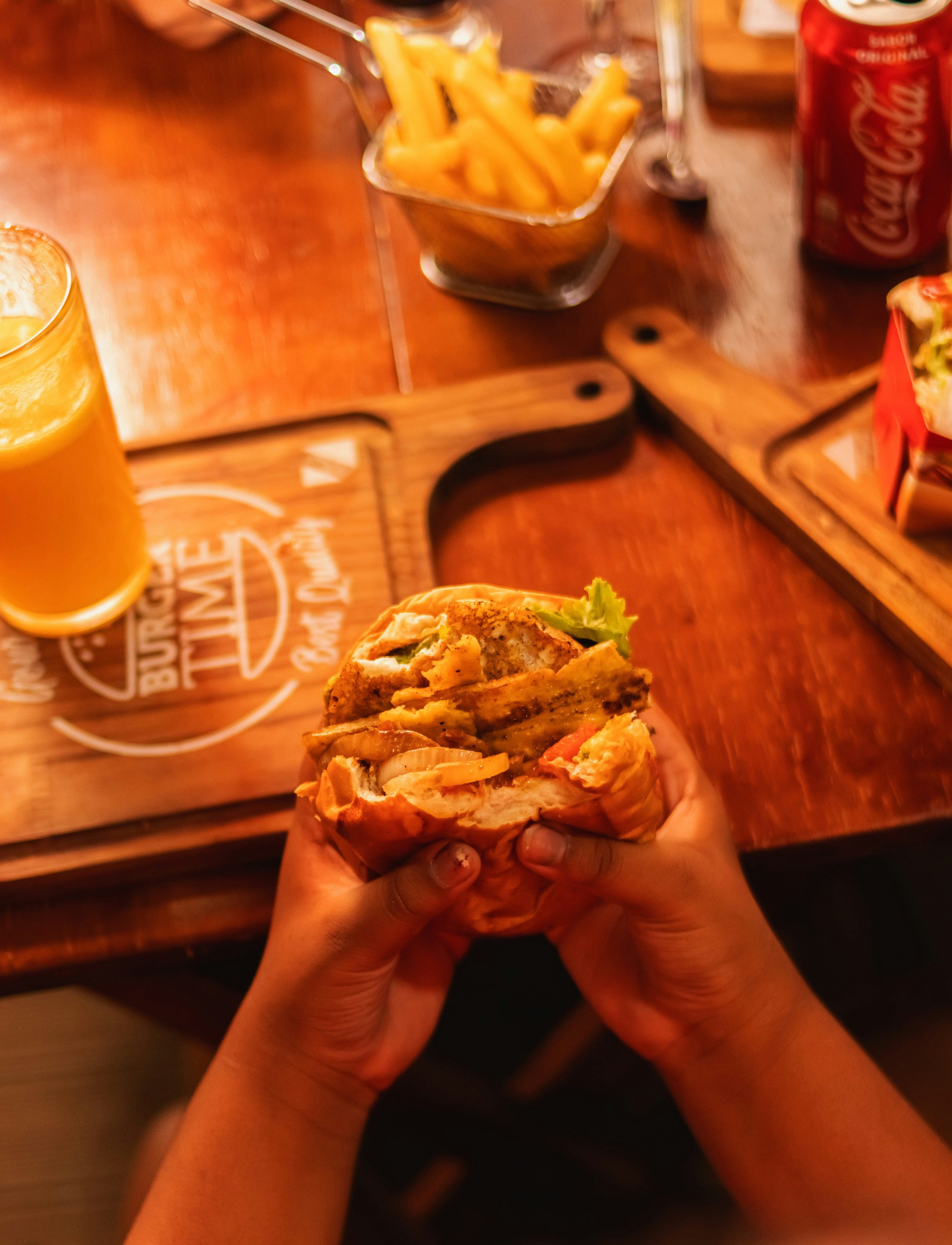 Hands gripping a towering burger filled with layers of ingredients, accompanied by a glass of orange drink and fries on a wooden table.
