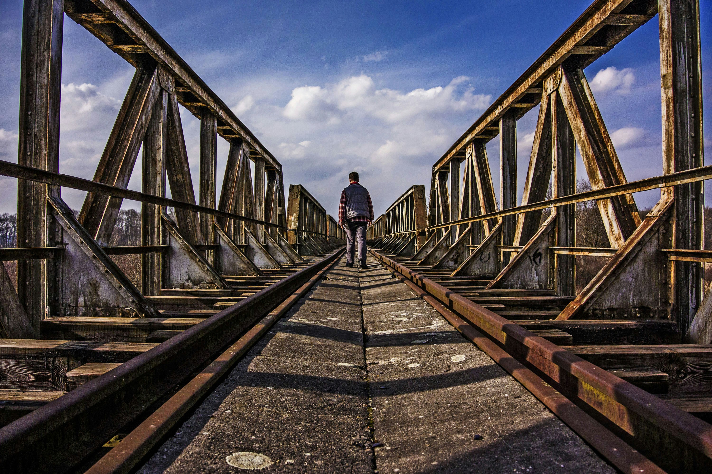 A solitary figure walks down an abandoned railway bridge, flanked by weathered wooden beams and a dramatic sky above.