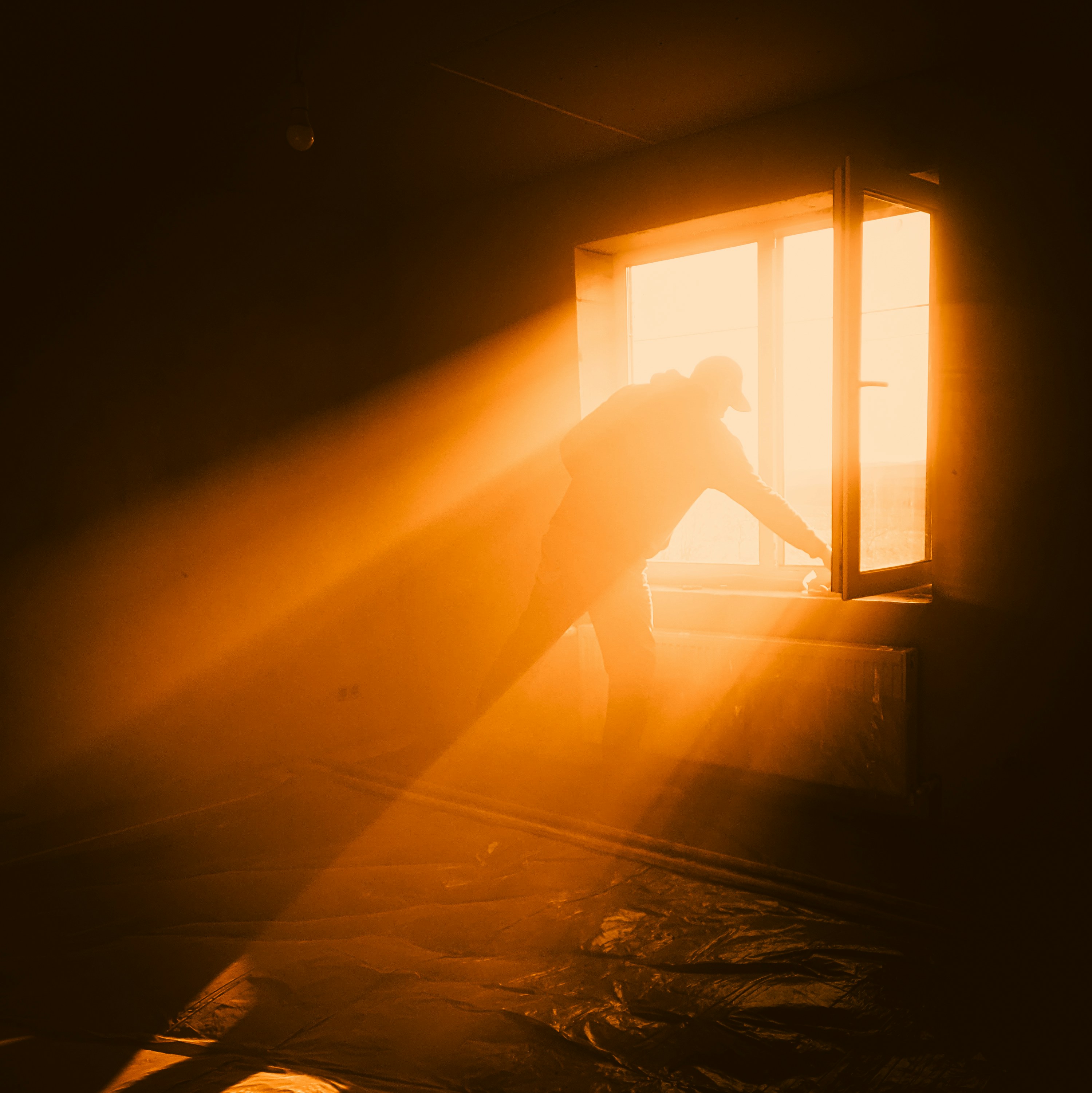 A silhouette of a person reaching out to a sunlit window, with warm beams of light streaming through the room. The atmosphere is filled with a soft, golden hue.