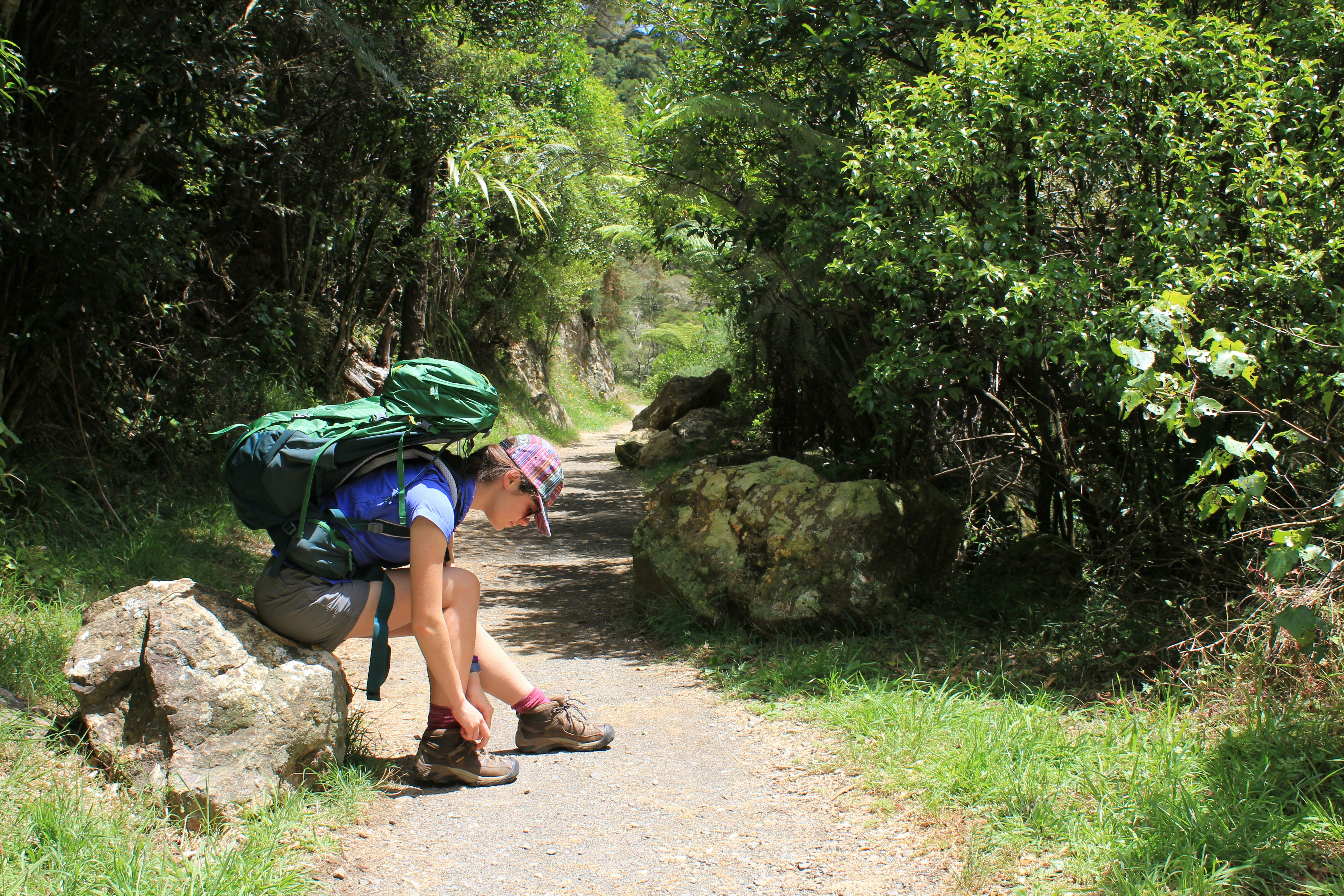 Hiker adjusting her boot laces on a forest trail surrounded by lush greenery and large rocks.