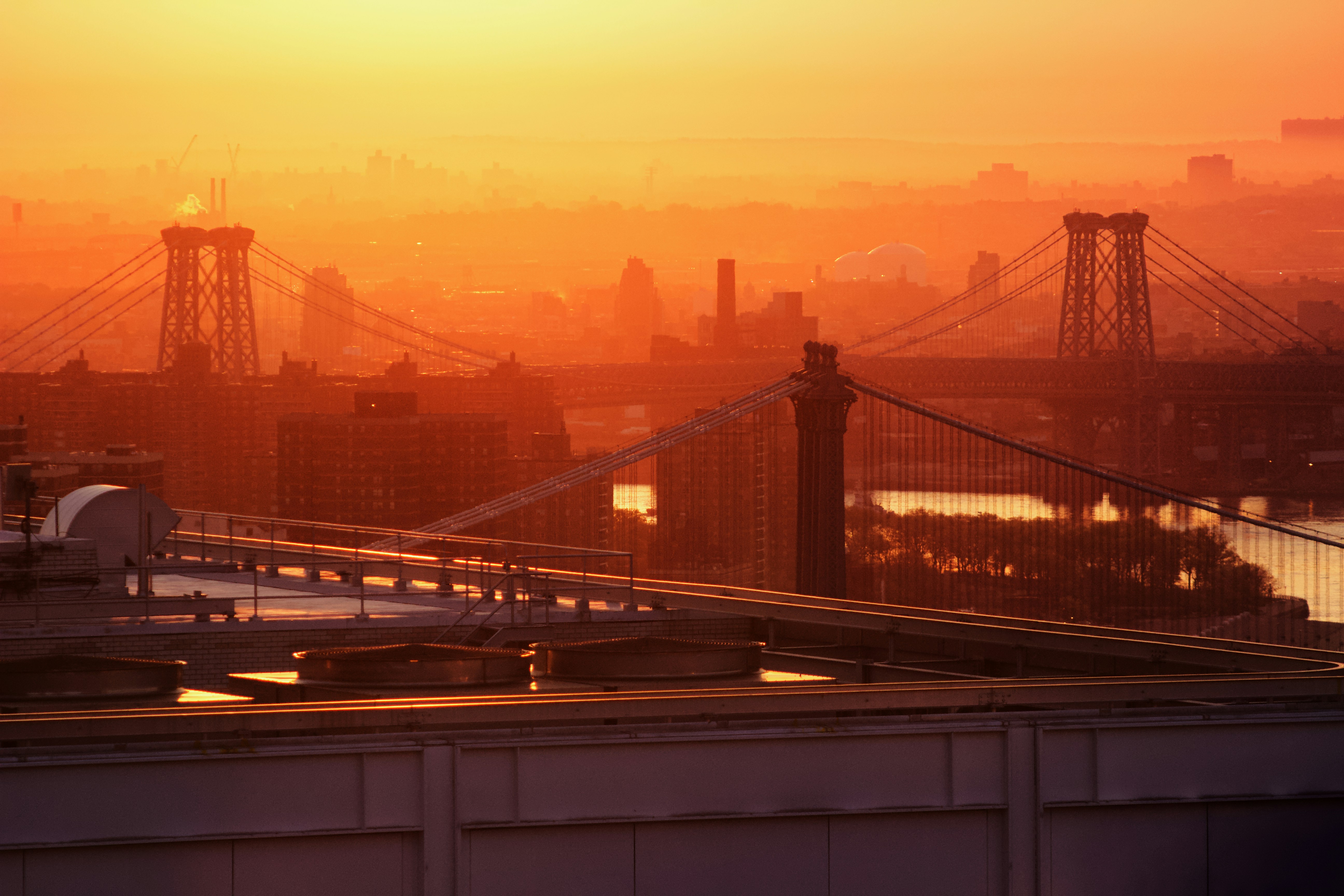 Golden sunrise illuminating the silhouettes of iconic bridges against a hazy skyline. The tranquil river reflects the warm hues of early morning.