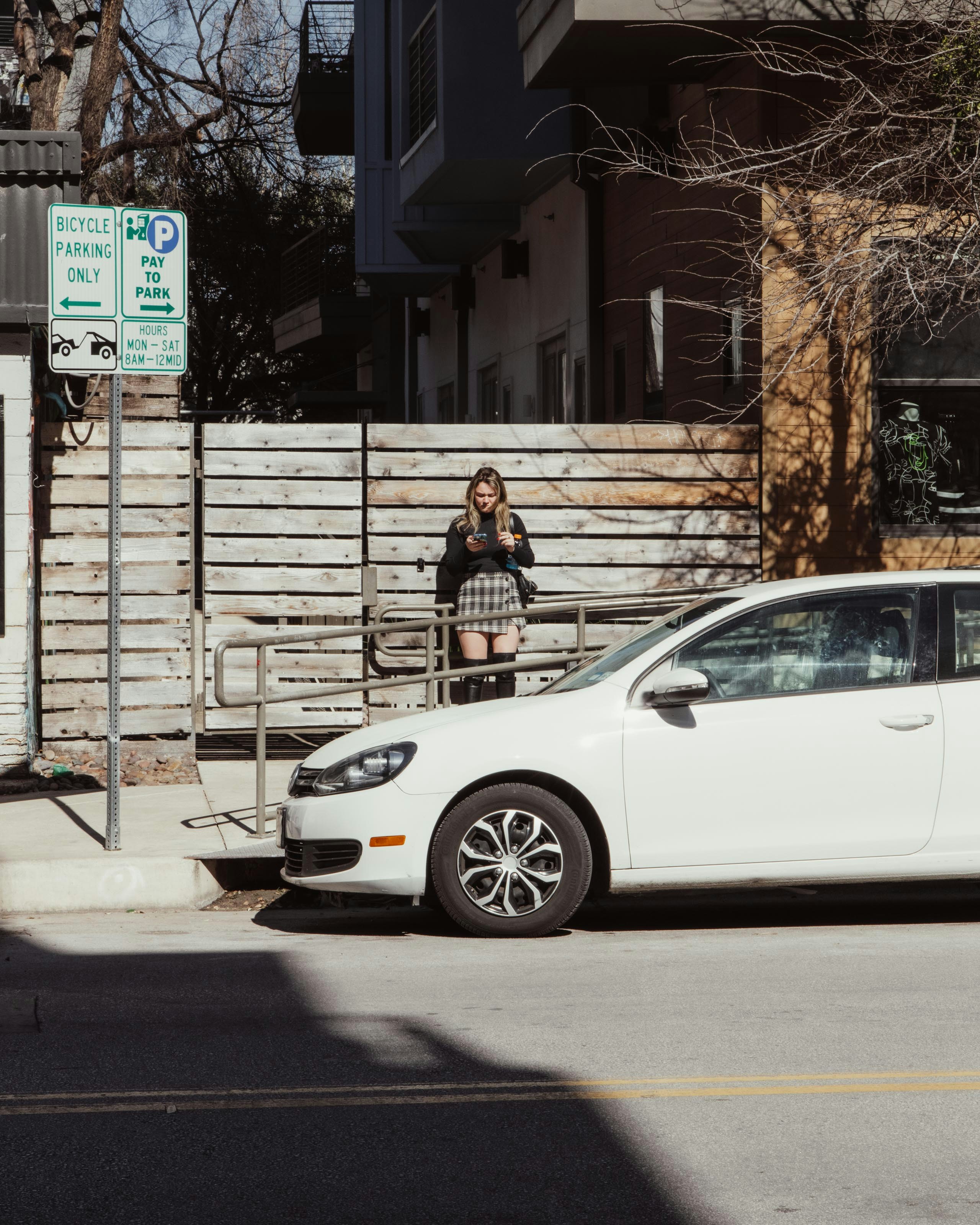 A young woman in a plaid skirt stands beside a white car on a city street, engrossed in her phone. The backdrop features a wooden fence and parking signs.