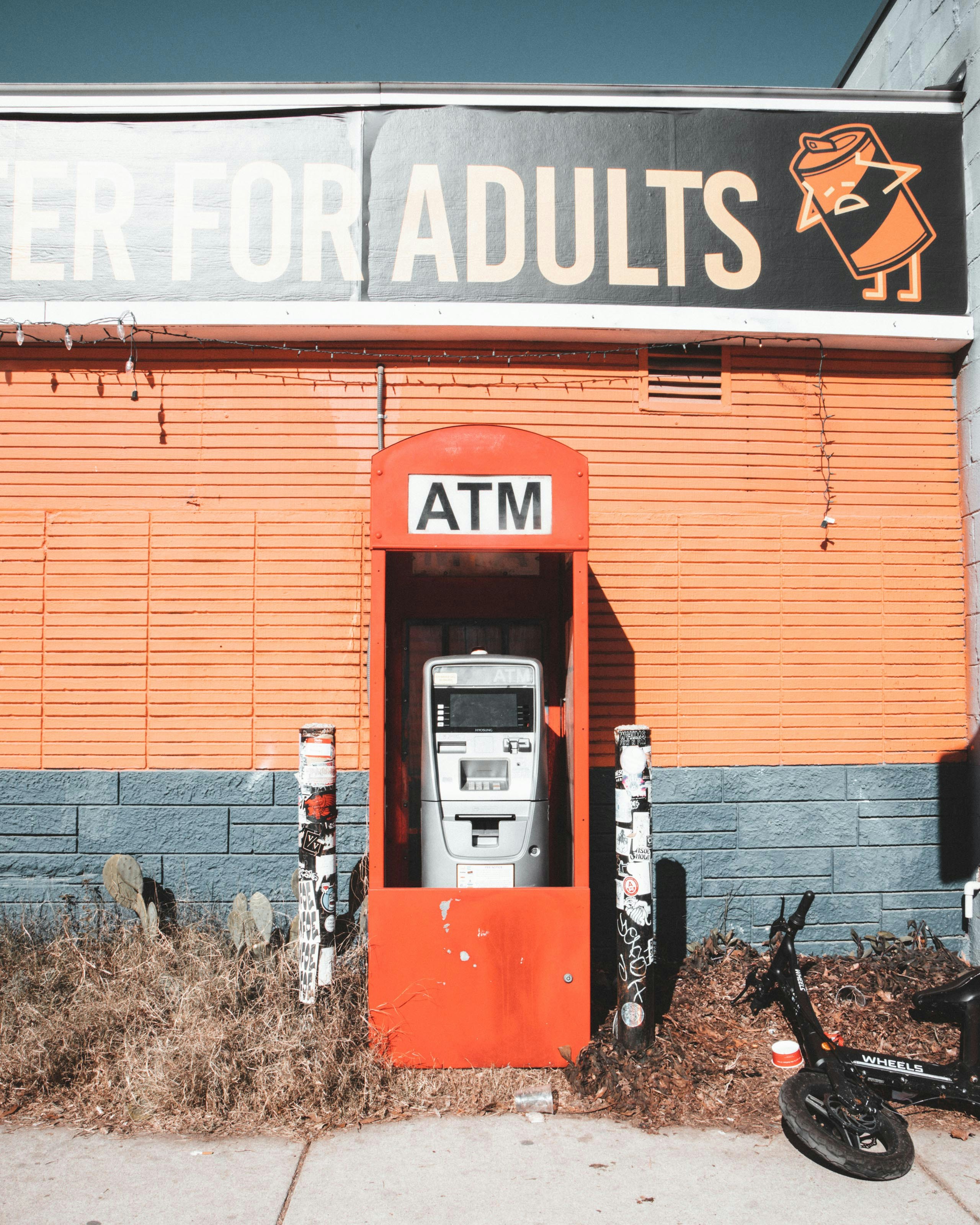 Bright orange ATM nestled against a colorful wall, surrounded by urban elements and overgrown vegetation.