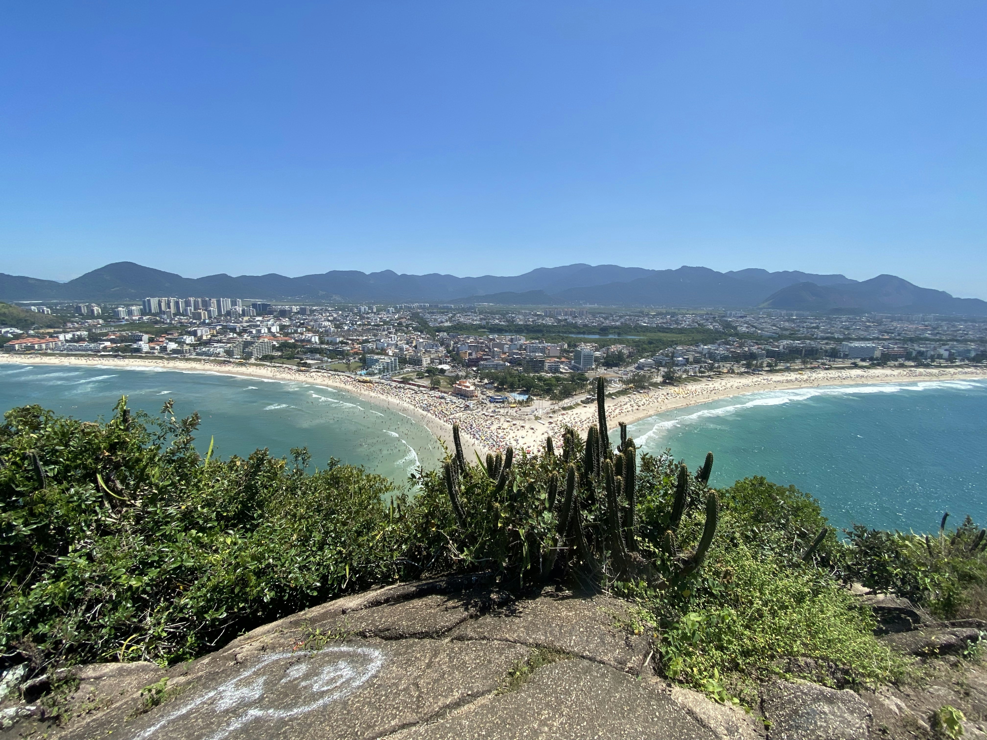 Aerial view of two curving beaches meeting at a lush, green promontory under a clear blue sky.