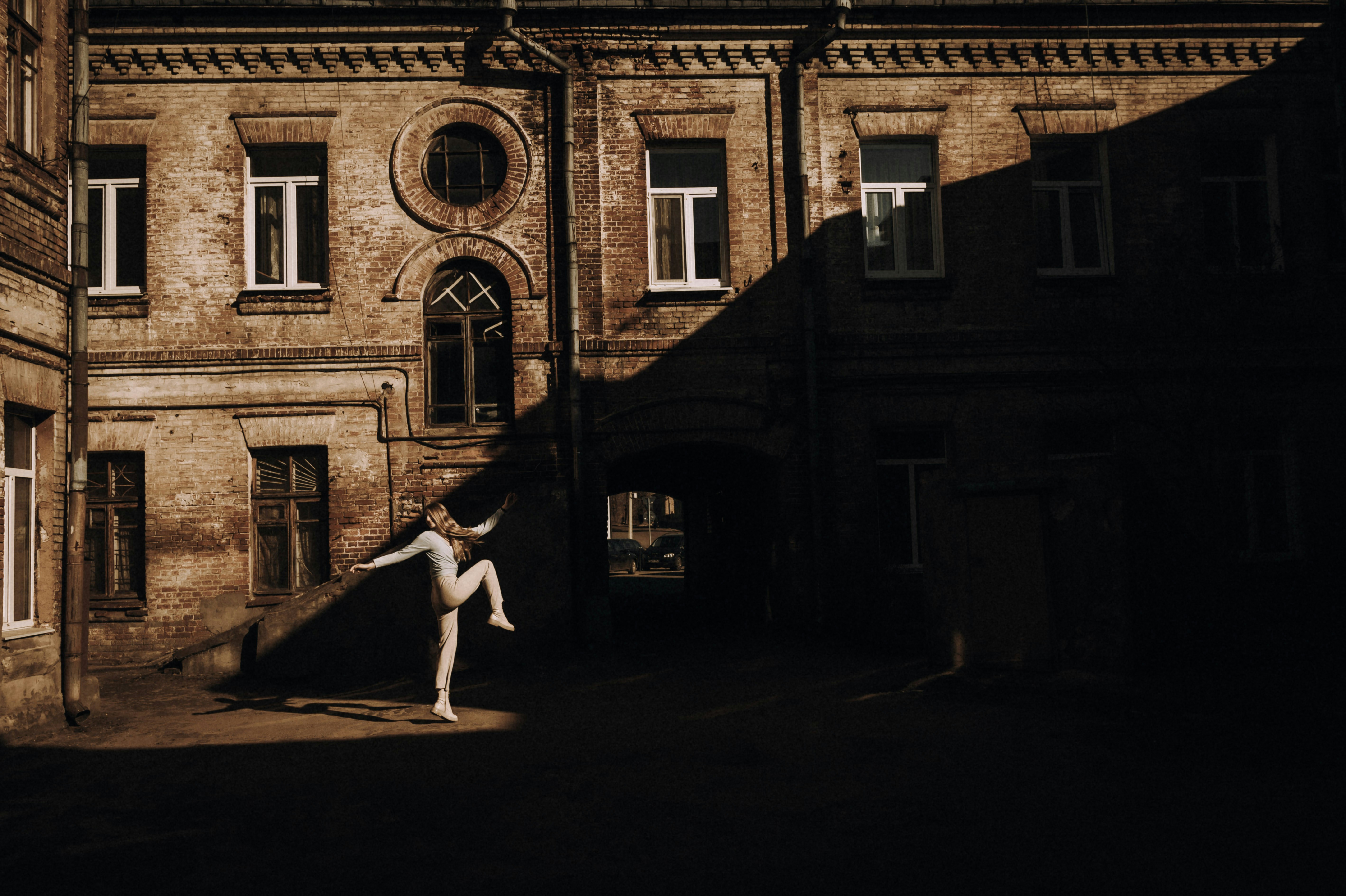 A dancer elegantly poses against a backdrop of an old brick building, with long shadows creating a dramatic contrast. The scene captures the interplay of light and movement.