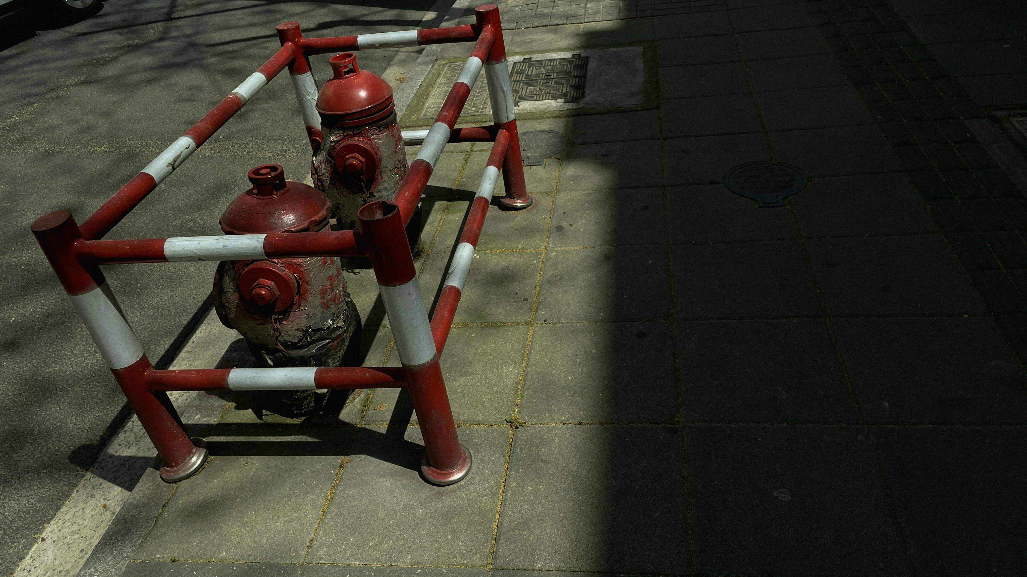 A trio of fire hydrants enclosed by a red and white barrier, casting shadows on a sunlit sidewalk.