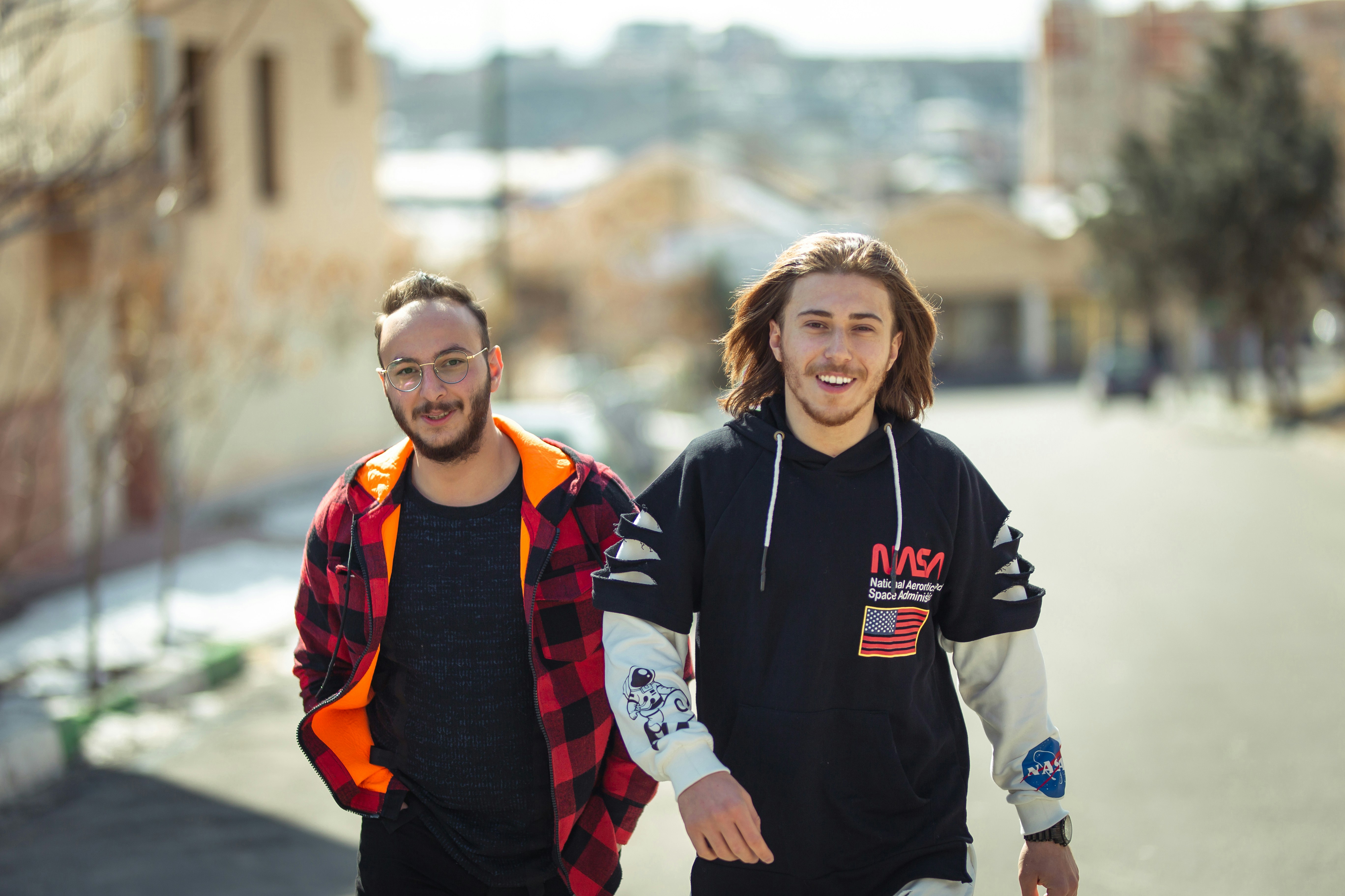 Two young men walk together down a sunlit street, showcasing their casual style and camaraderie.