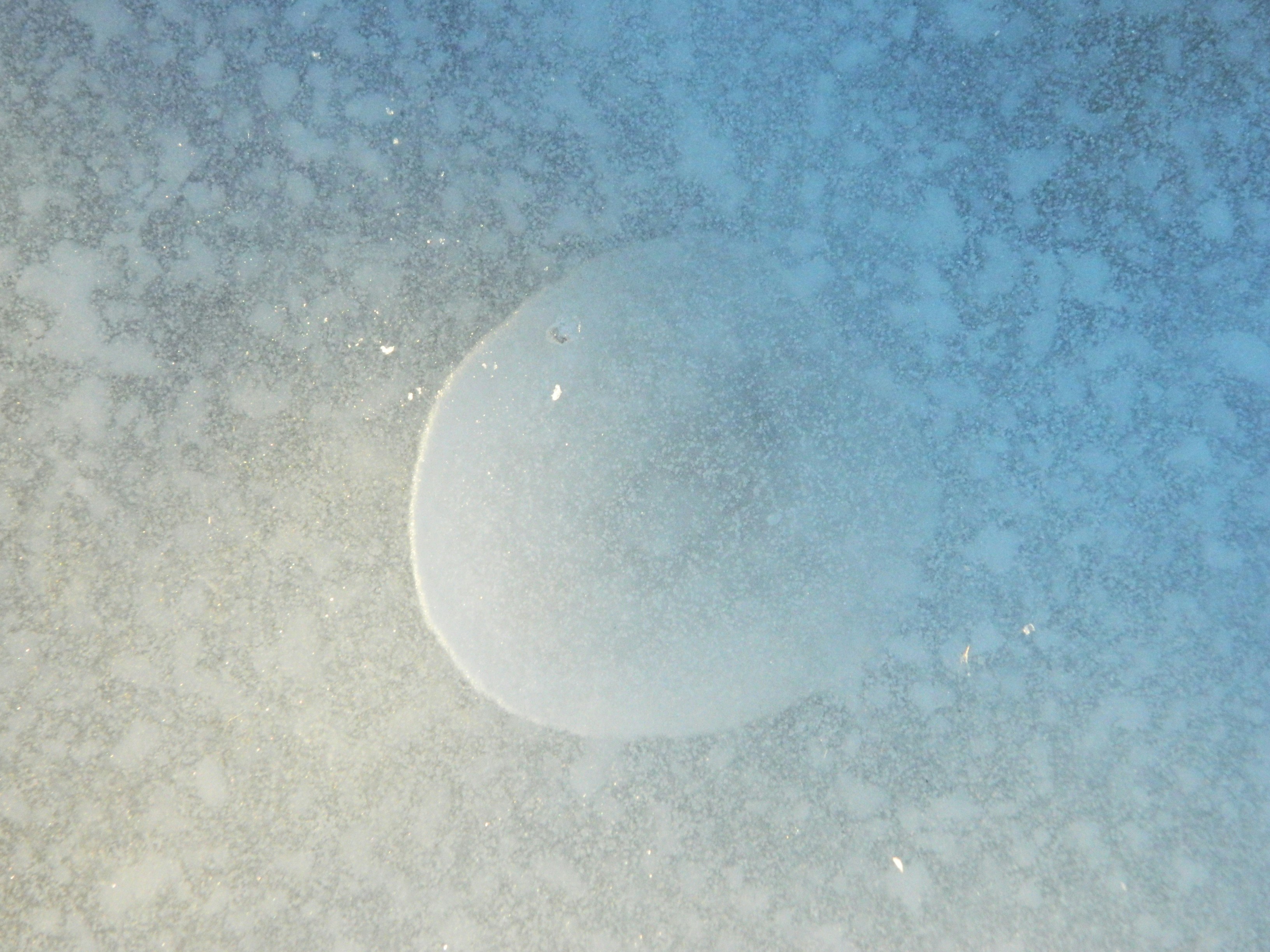A circular frozen bubble rests on a textured icy surface, capturing the delicate interplay of frost and light. The scene evokes a serene winter atmosphere.