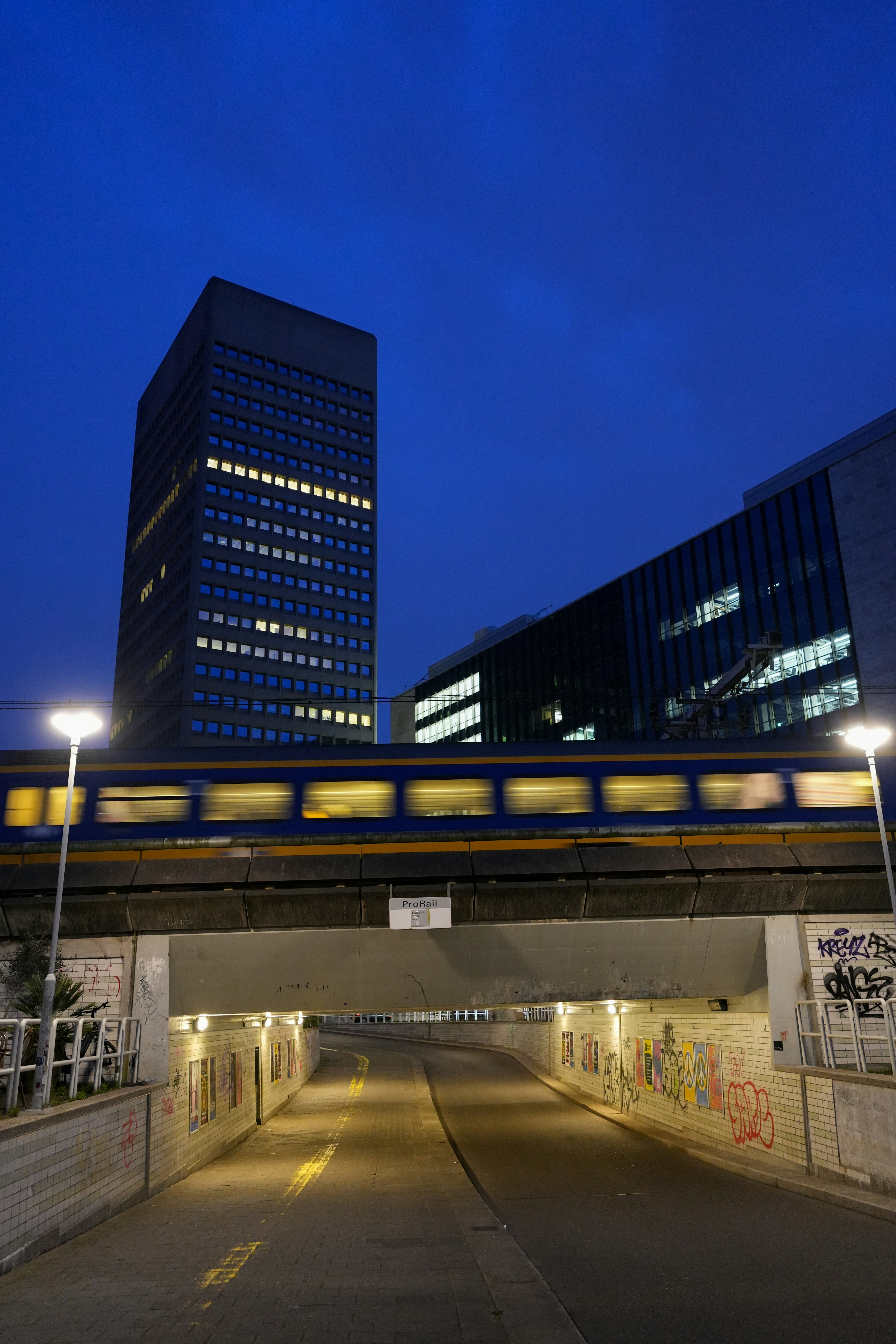 A train speeds through an urban underpass illuminated by streetlights, with modern buildings towering in the background. Graffiti adorns the walls, contrasting with the sleek architecture.