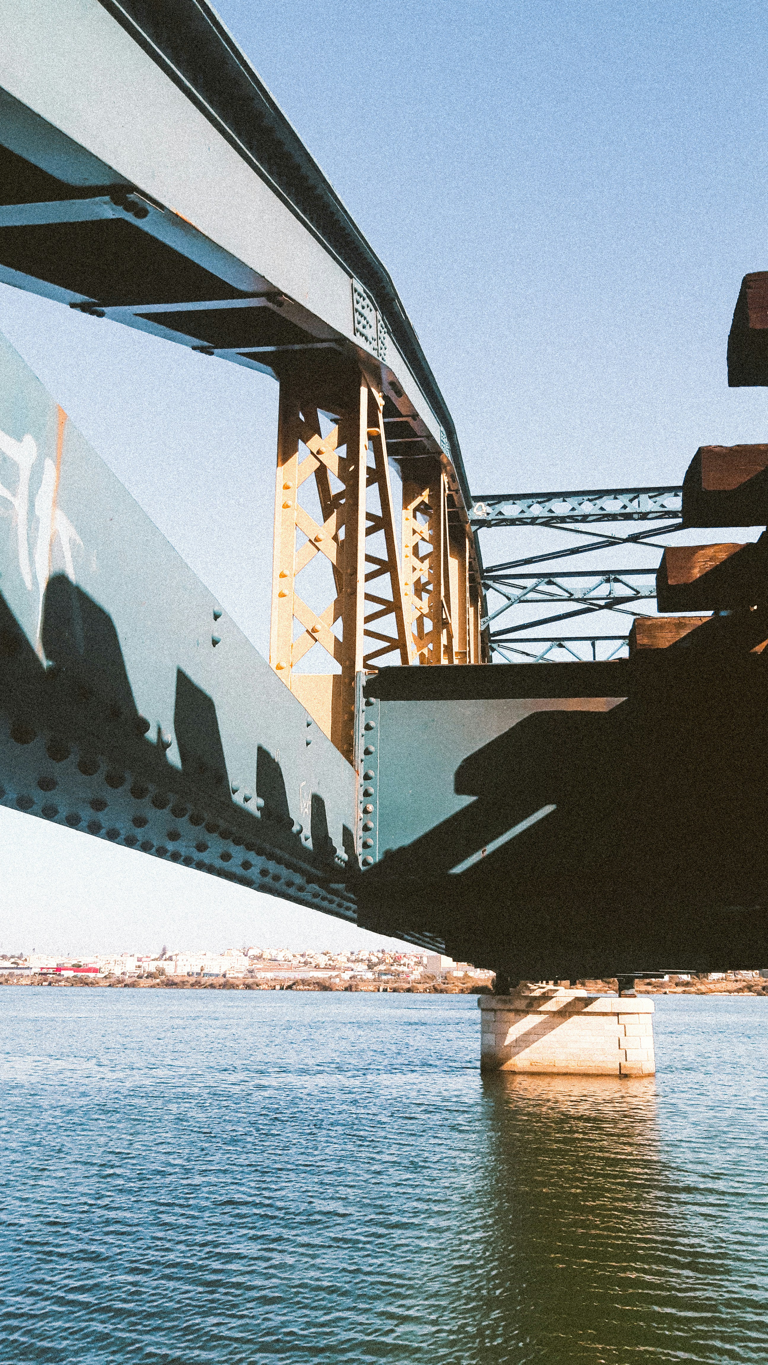 Steel undercarriage of a bridge extends over calm water under a clear sky. Sunlit rivets and cross-braces form graphic geometry along the span.