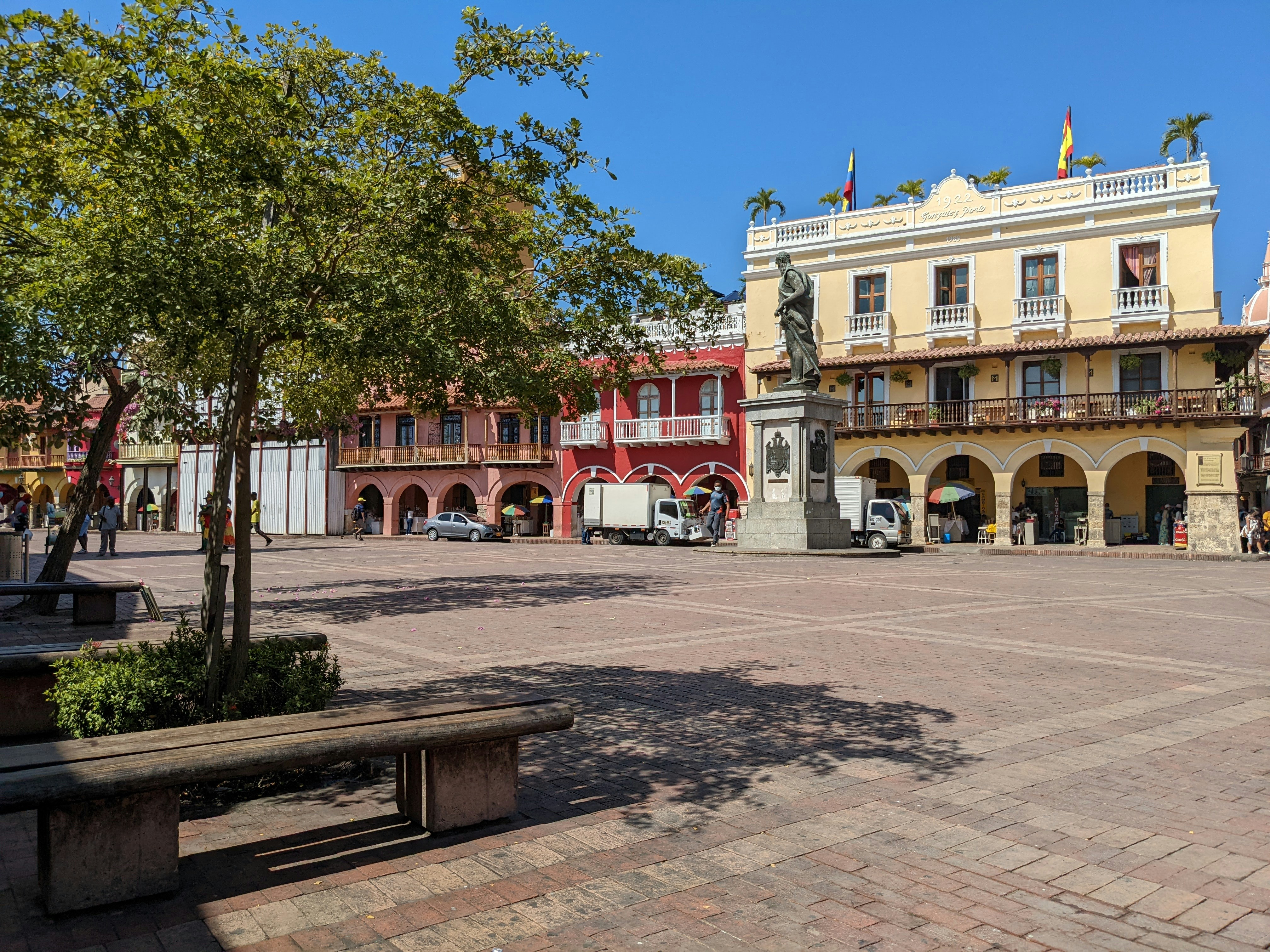Tree-lined plaza with colorful buildings and clear blue sky.