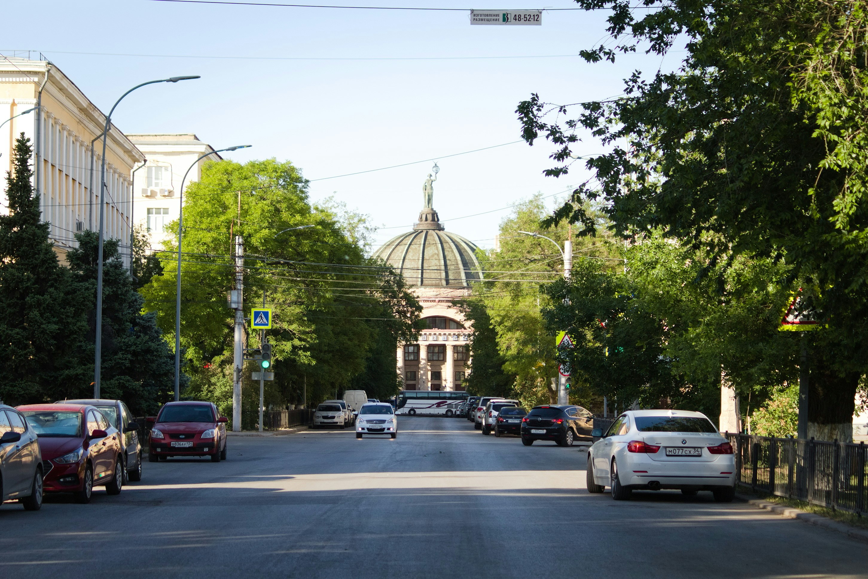 Cars line a tree-shaded street leading to a domed building under a clear sky.