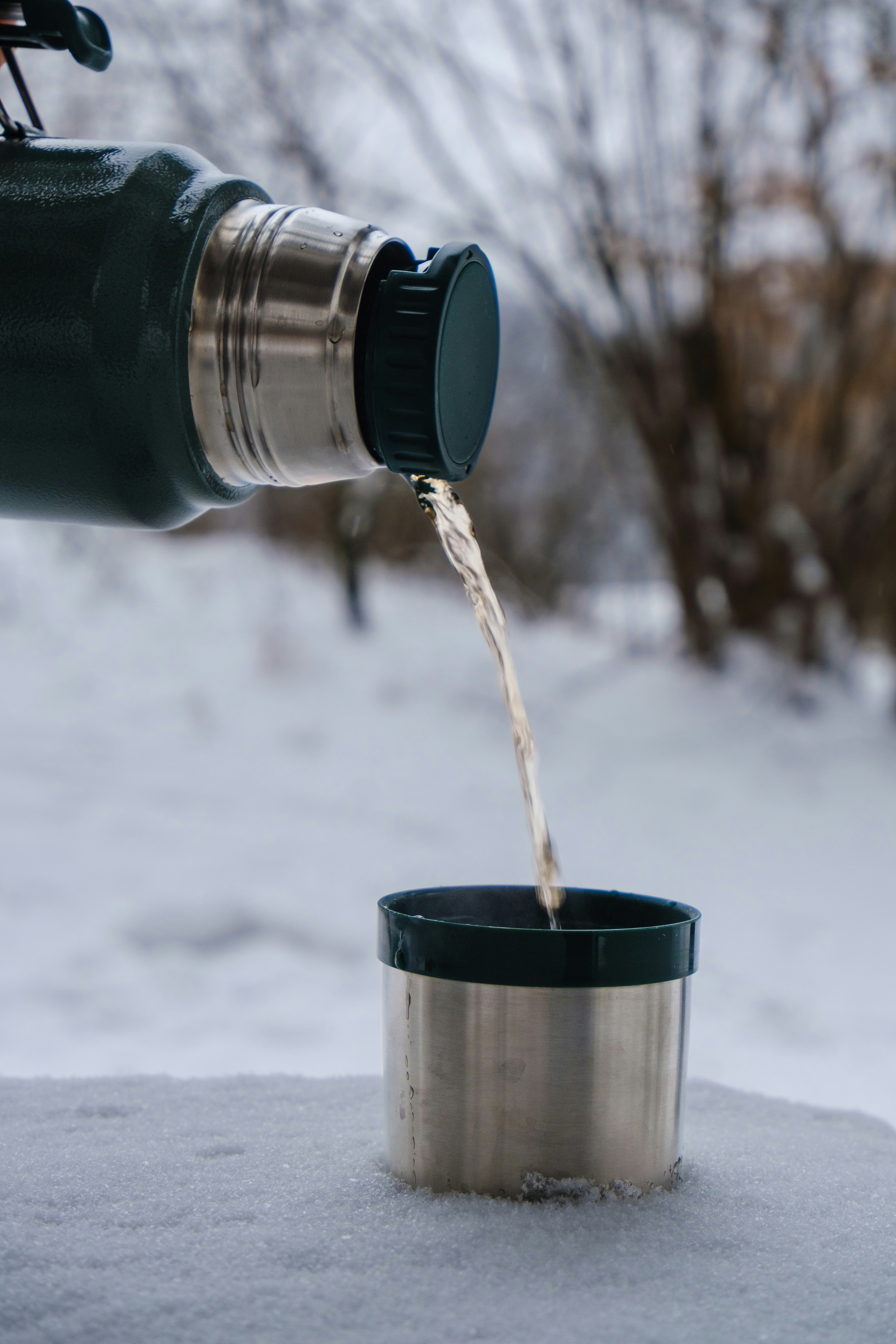 Hot beverage pouring from a thermos into a stainless steel cup, surrounded by a snowy landscape.