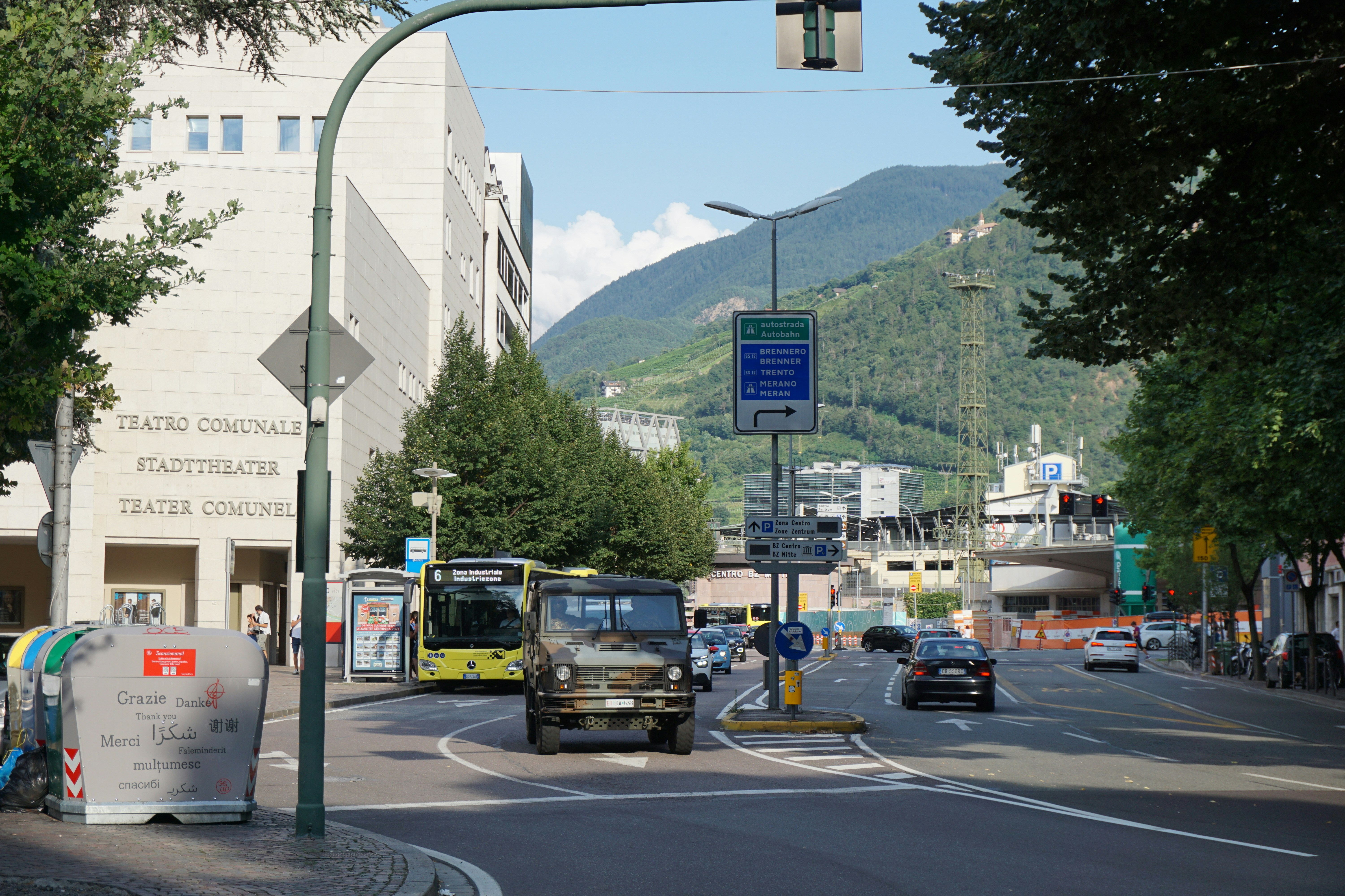A bustling city street featuring a mix of vehicles and pedestrians near a modern theater, framed by lush greenery and distant mountains.