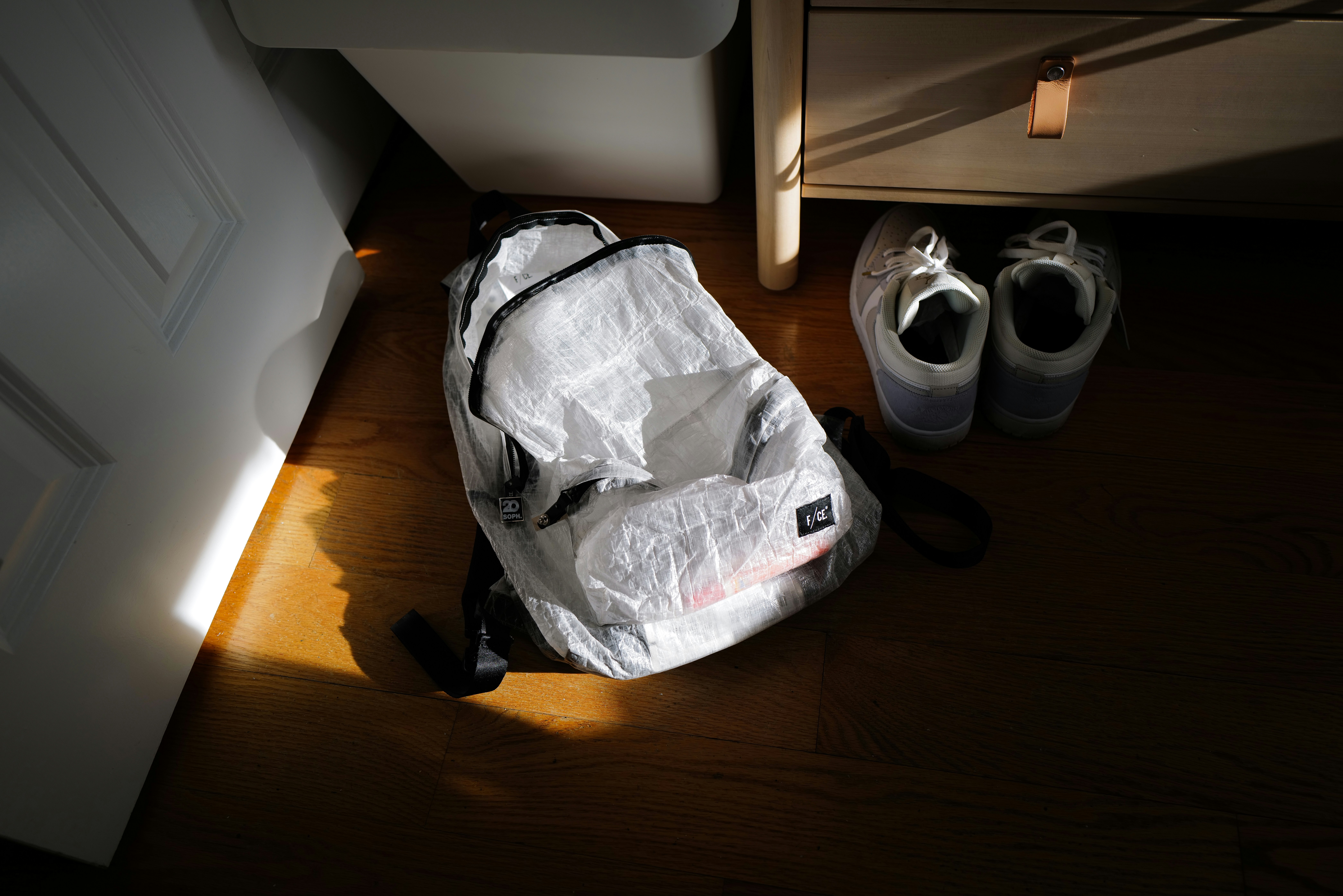 Sunlit Corner Loadout: Crumpled Backpack Resting by Sneakers on Warm Hardwood
