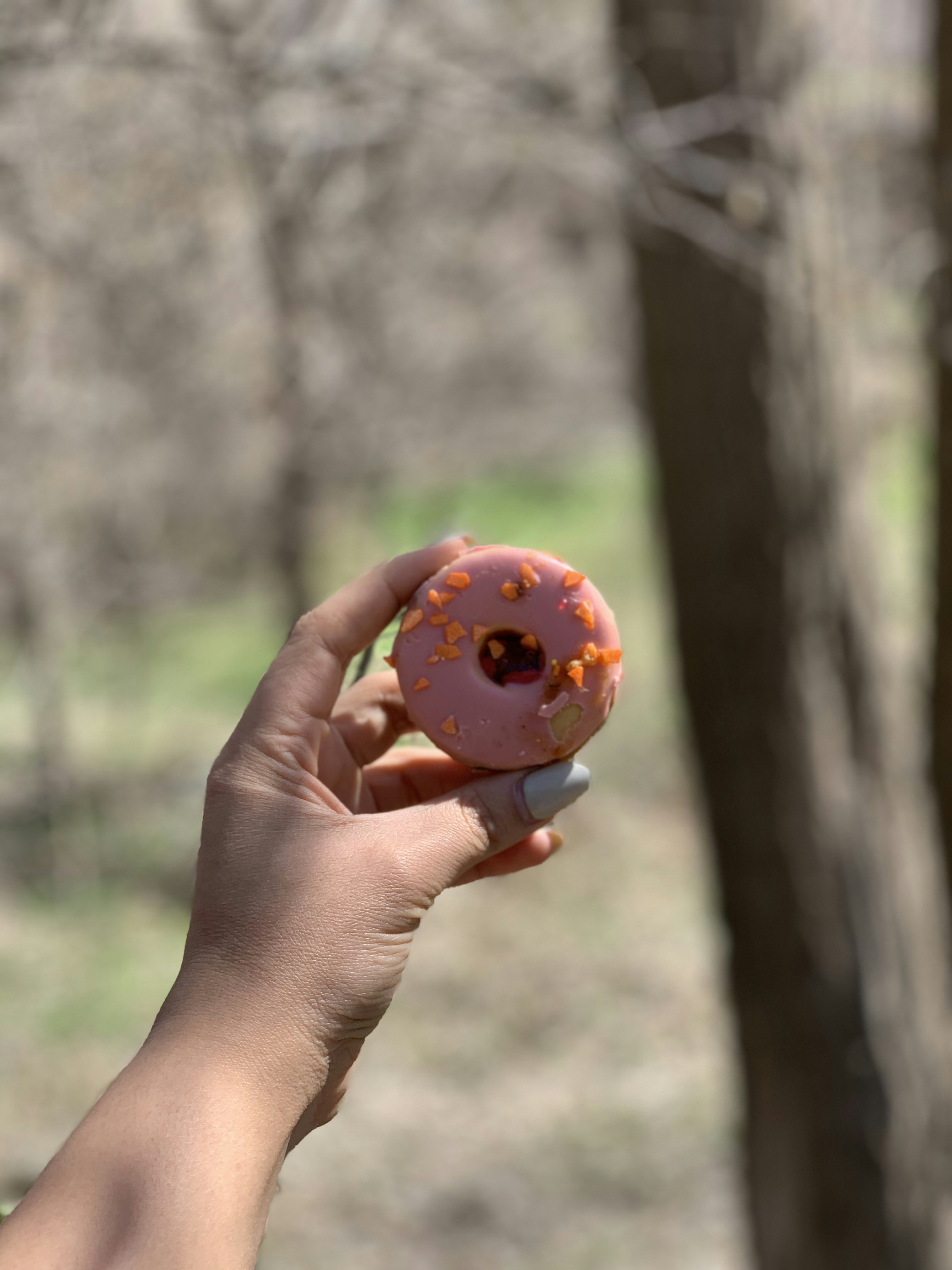 A hand holds a pink frosted donut with orange sprinkles against a blurred natural background. The focus is on the donut, showcasing its vibrant color.