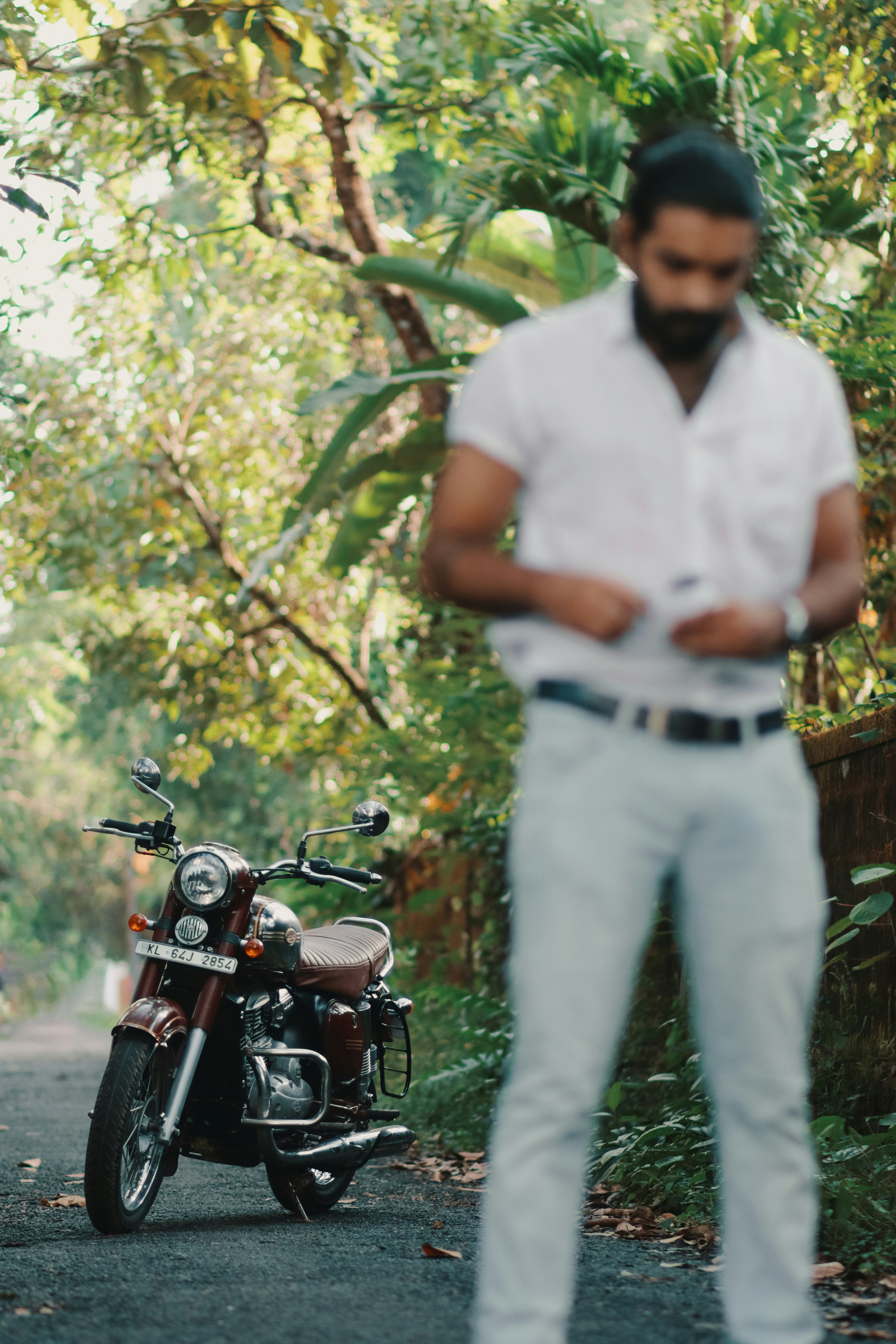 A man stands in the foreground, adjusting his phone, while a vintage motorcycle rests on a serene, tree-lined road in the background.