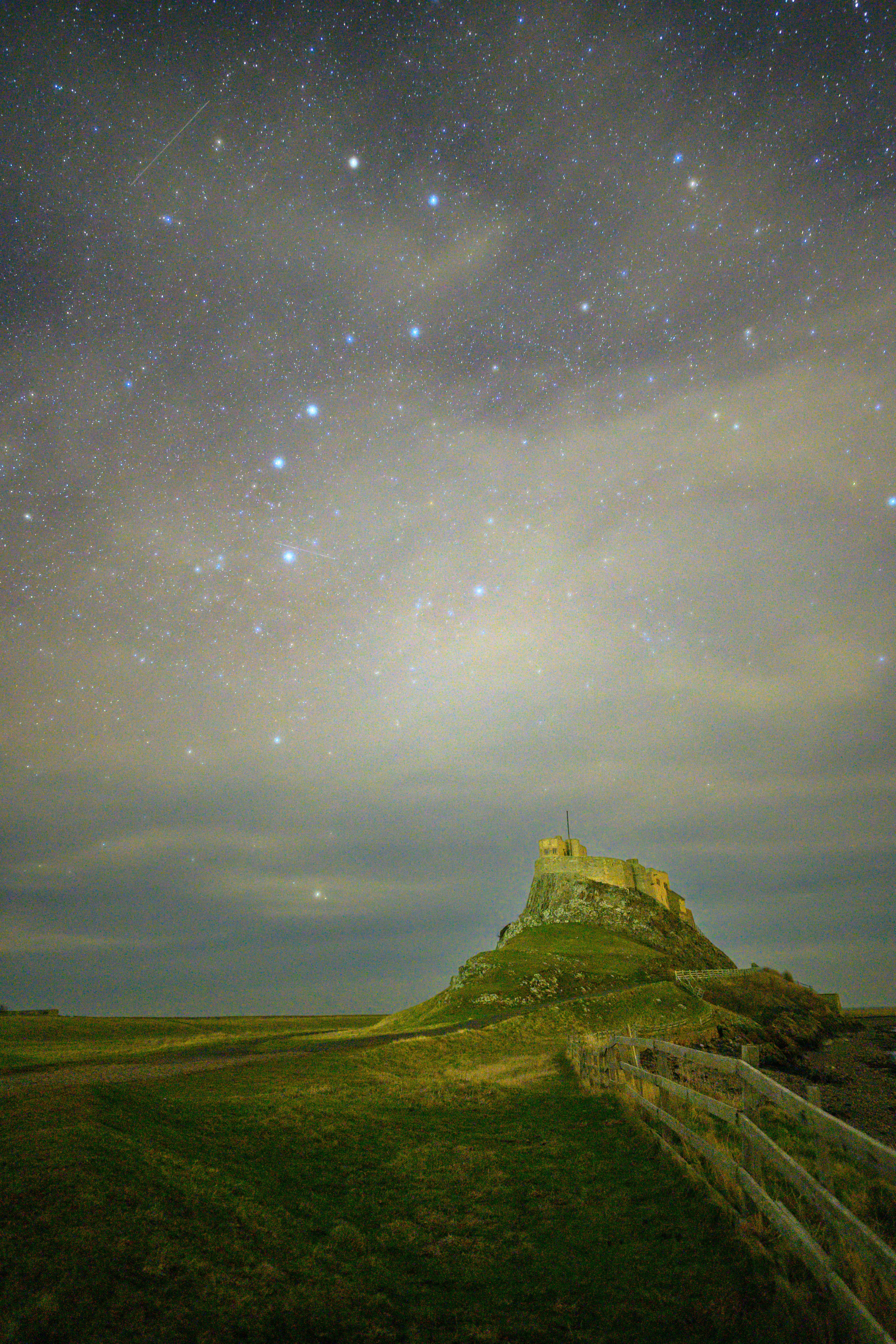 Historic castle perched on a grassy hill under a star-filled sky, with wispy clouds adding depth to the night scene.