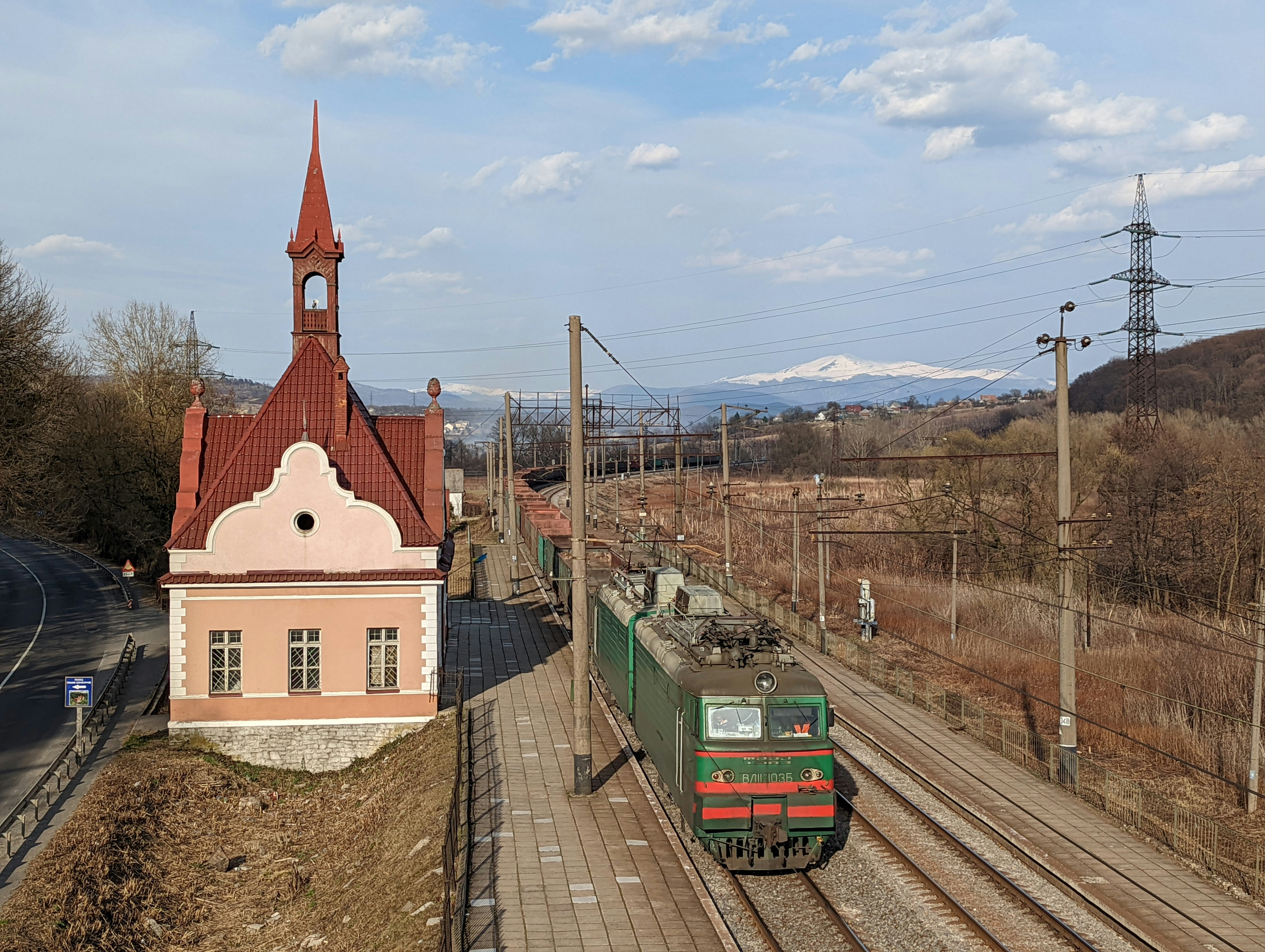 Historic station building alongside a railway track as a green train approaches, set against a backdrop of distant mountains.