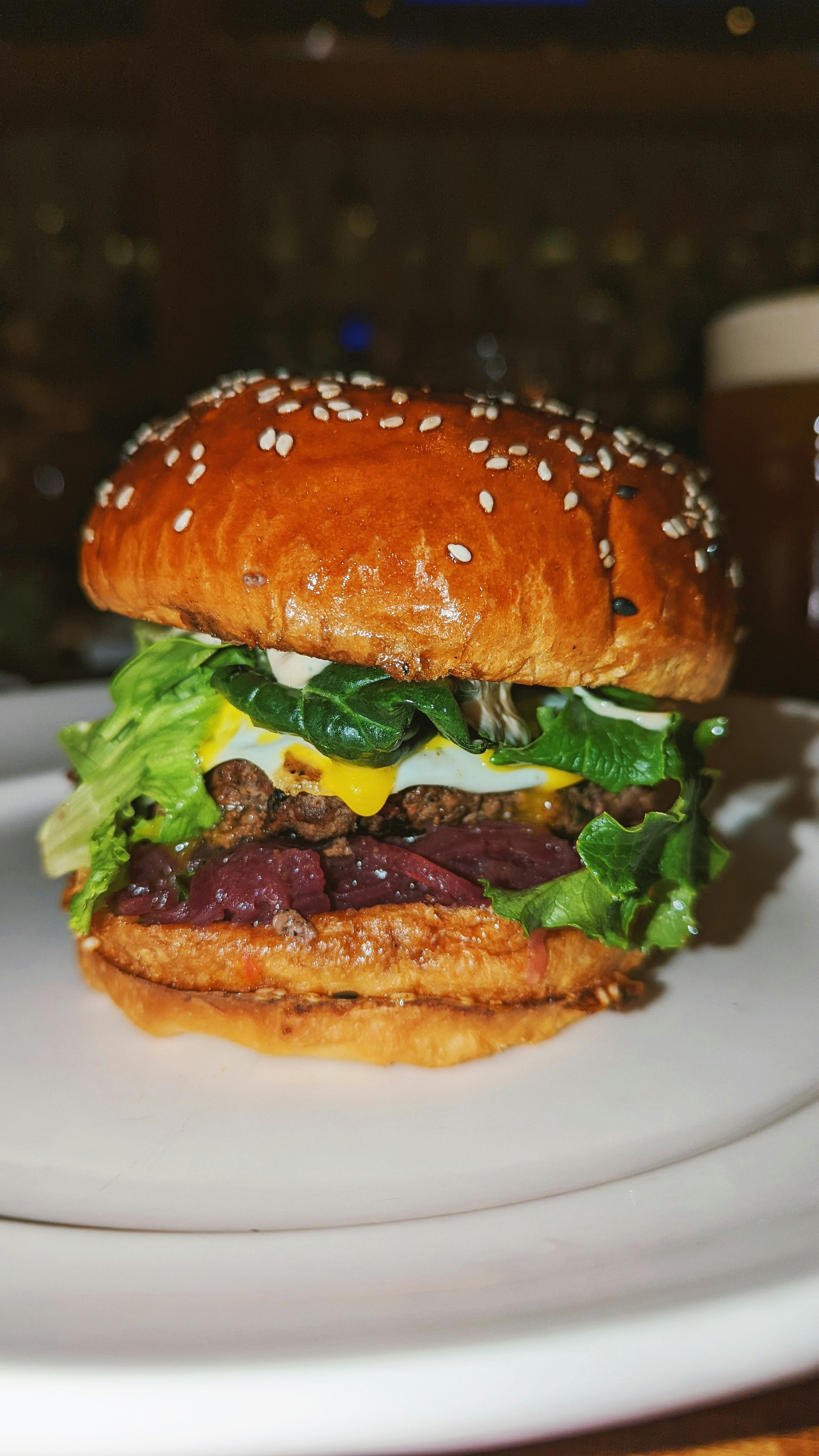 Close-up of a sesame-seed bun burger with lettuce, cheese, and a beef patty on a white plate, set against a warm, dim cafe backdrop.