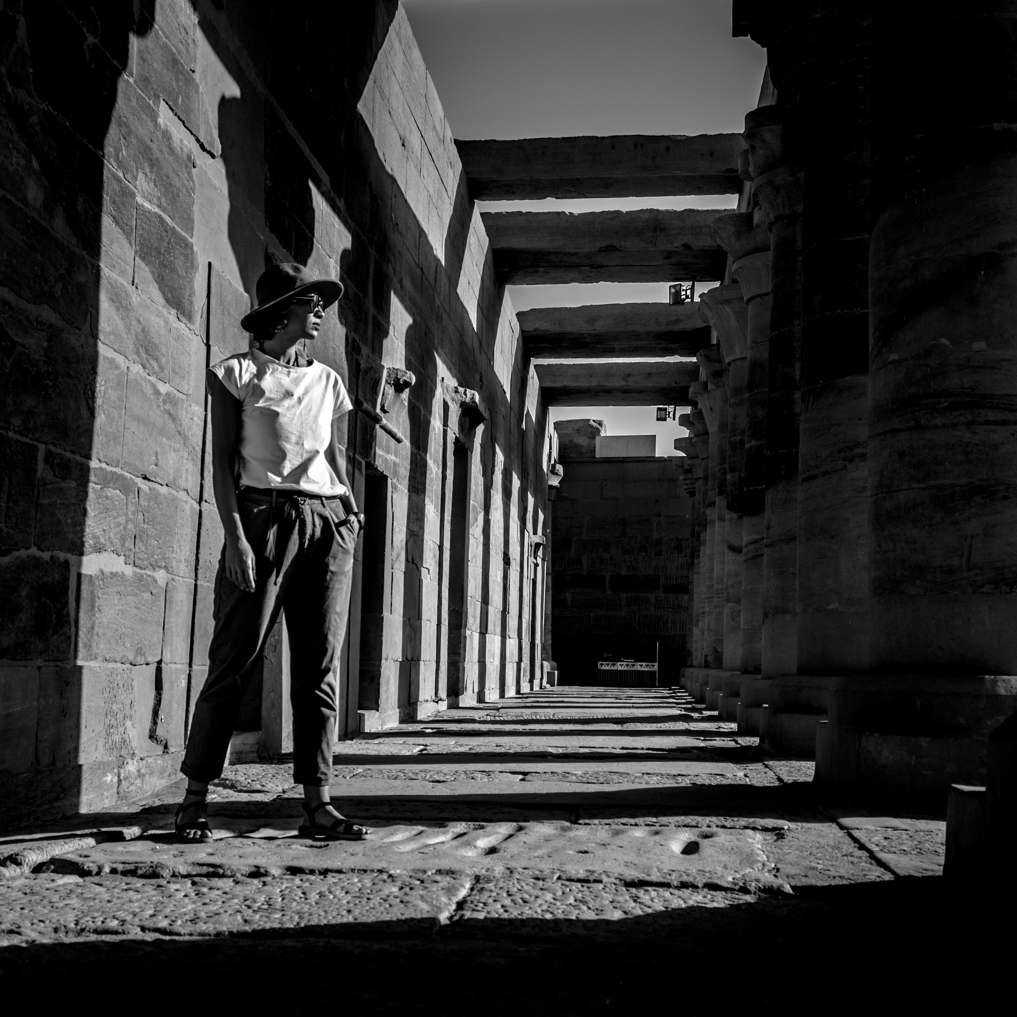 A figure stands in a sunlit corridor of ancient stone architecture, casting long shadows in a monochrome setting.