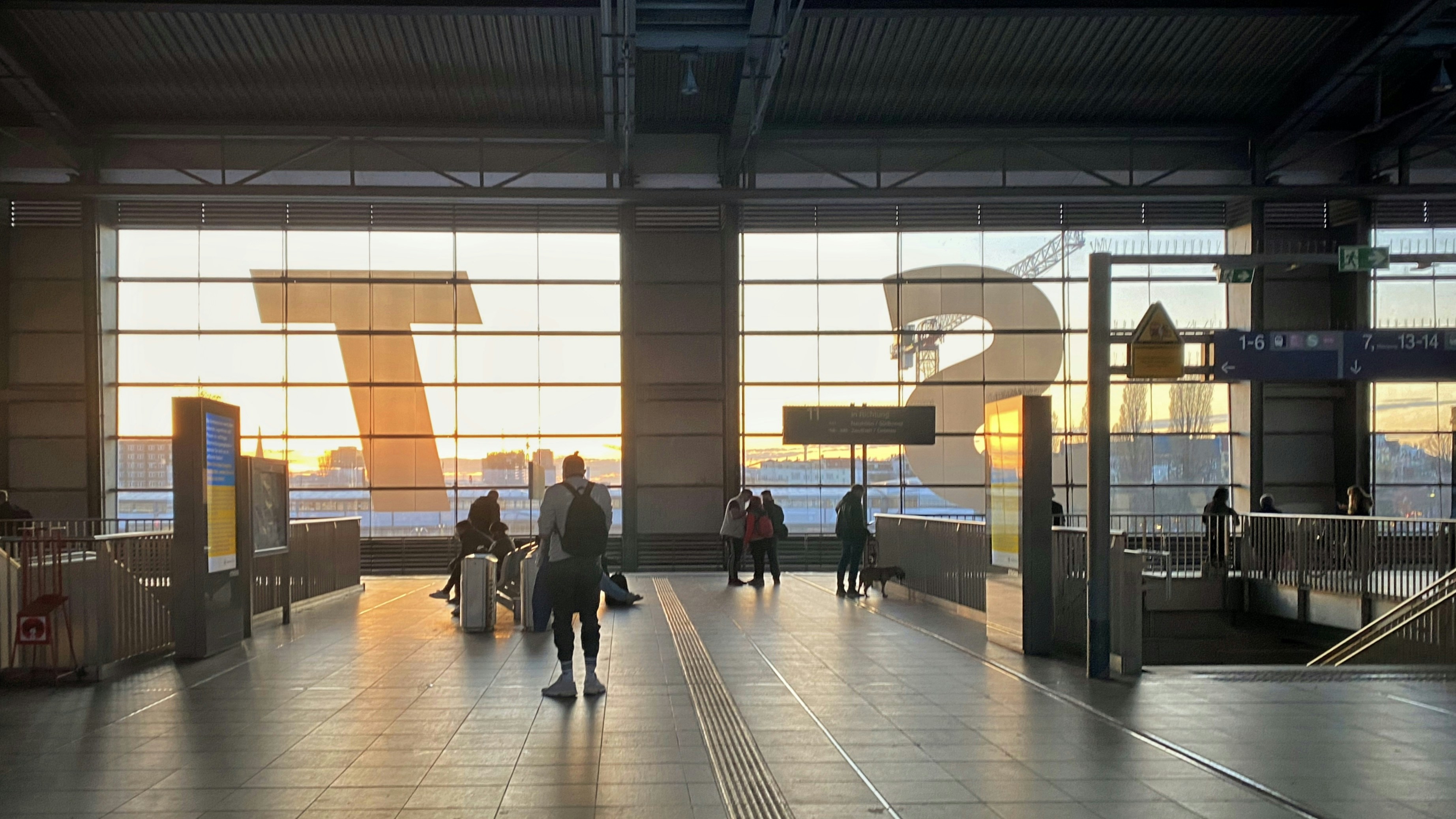 Silhouetted figures move through a train station as the sun sets, casting warm light through large glass windows. The letters 'S T' are prominently displayed, hinting at the station's name.