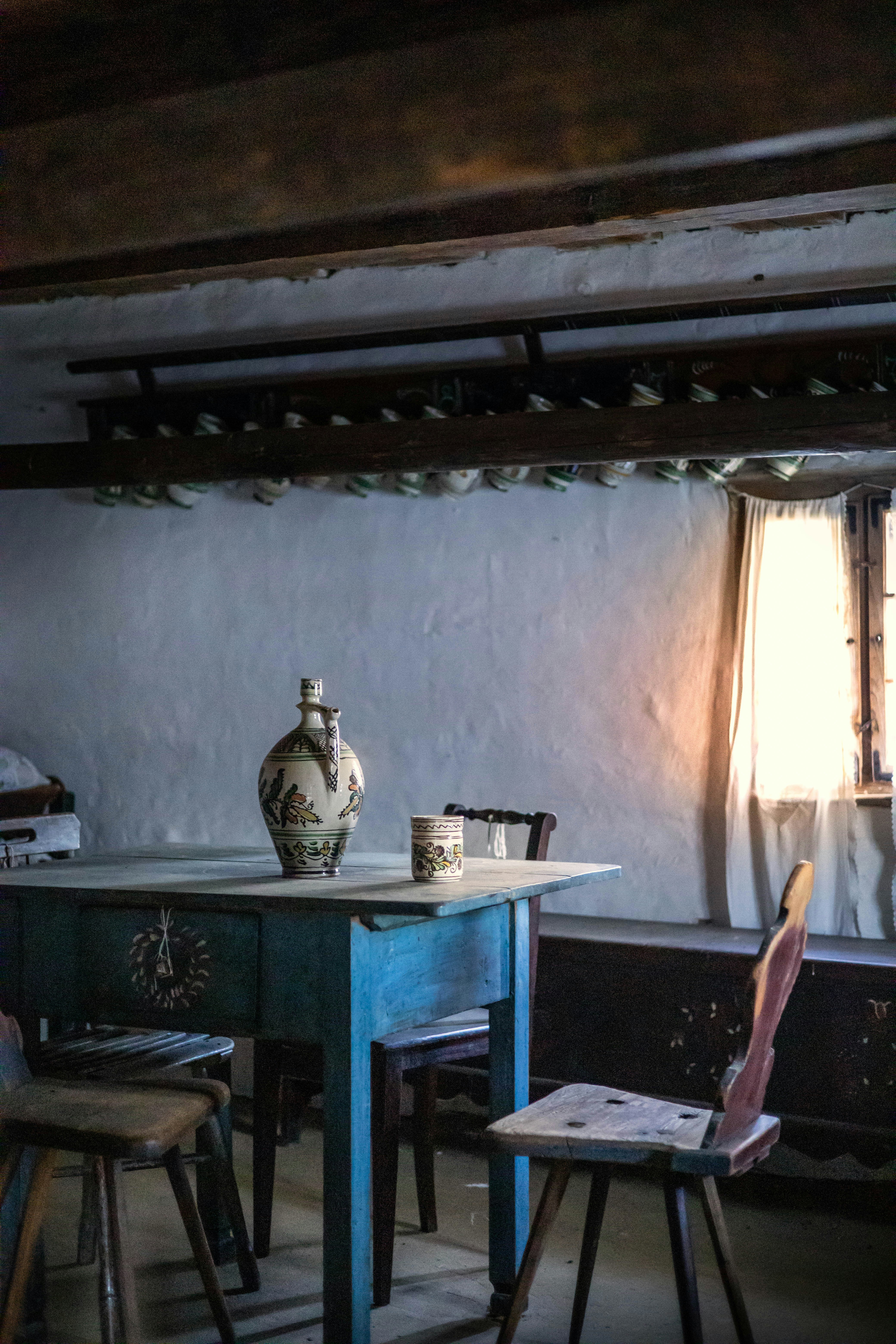 A vintage blue table adorned with a decorative vase and a mug, illuminated by soft natural light streaming through a window in a cozy room.