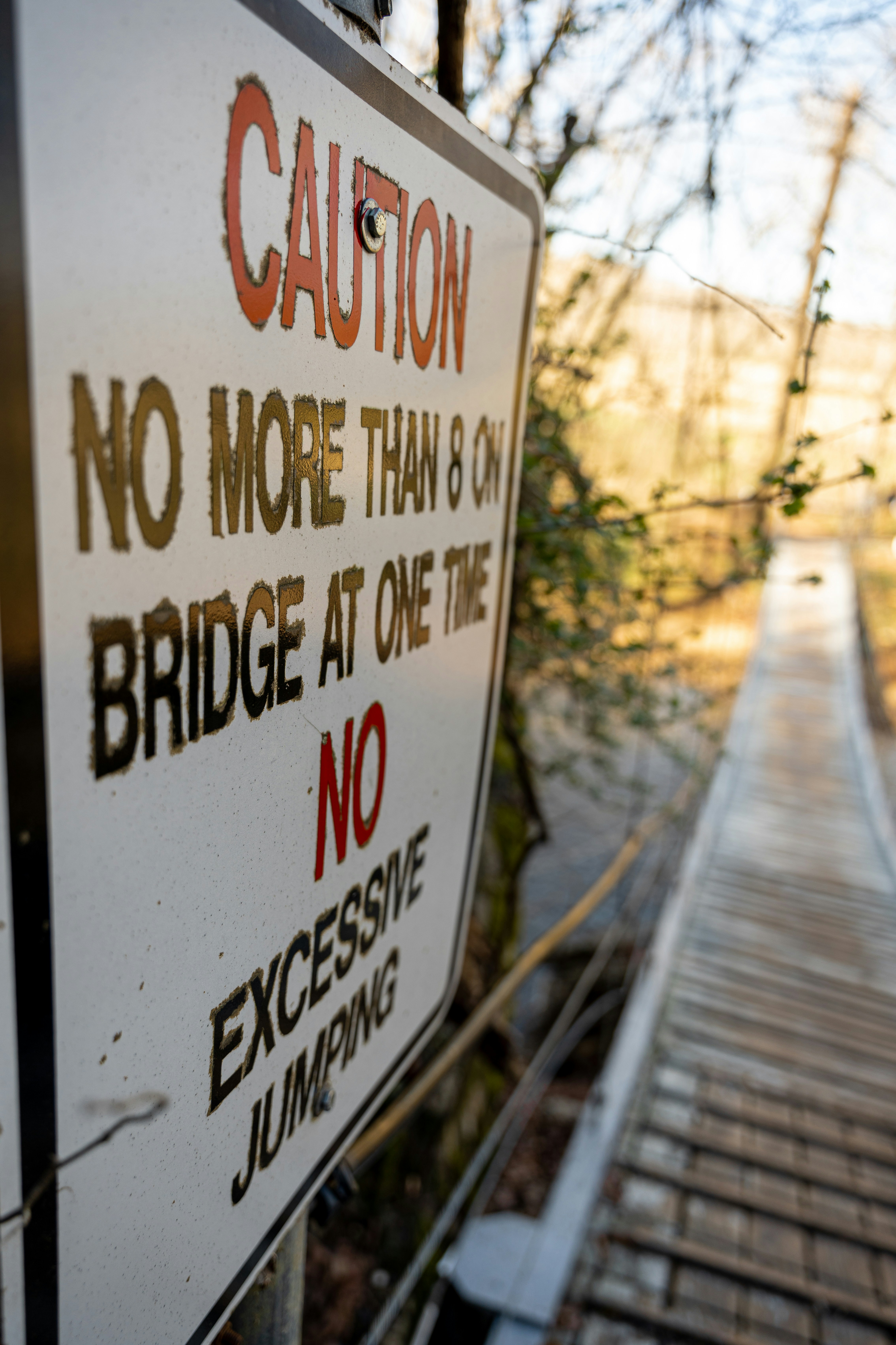 Caution sign on a bridge warning against excessive jumping, with a wooden bridge leading into the background.