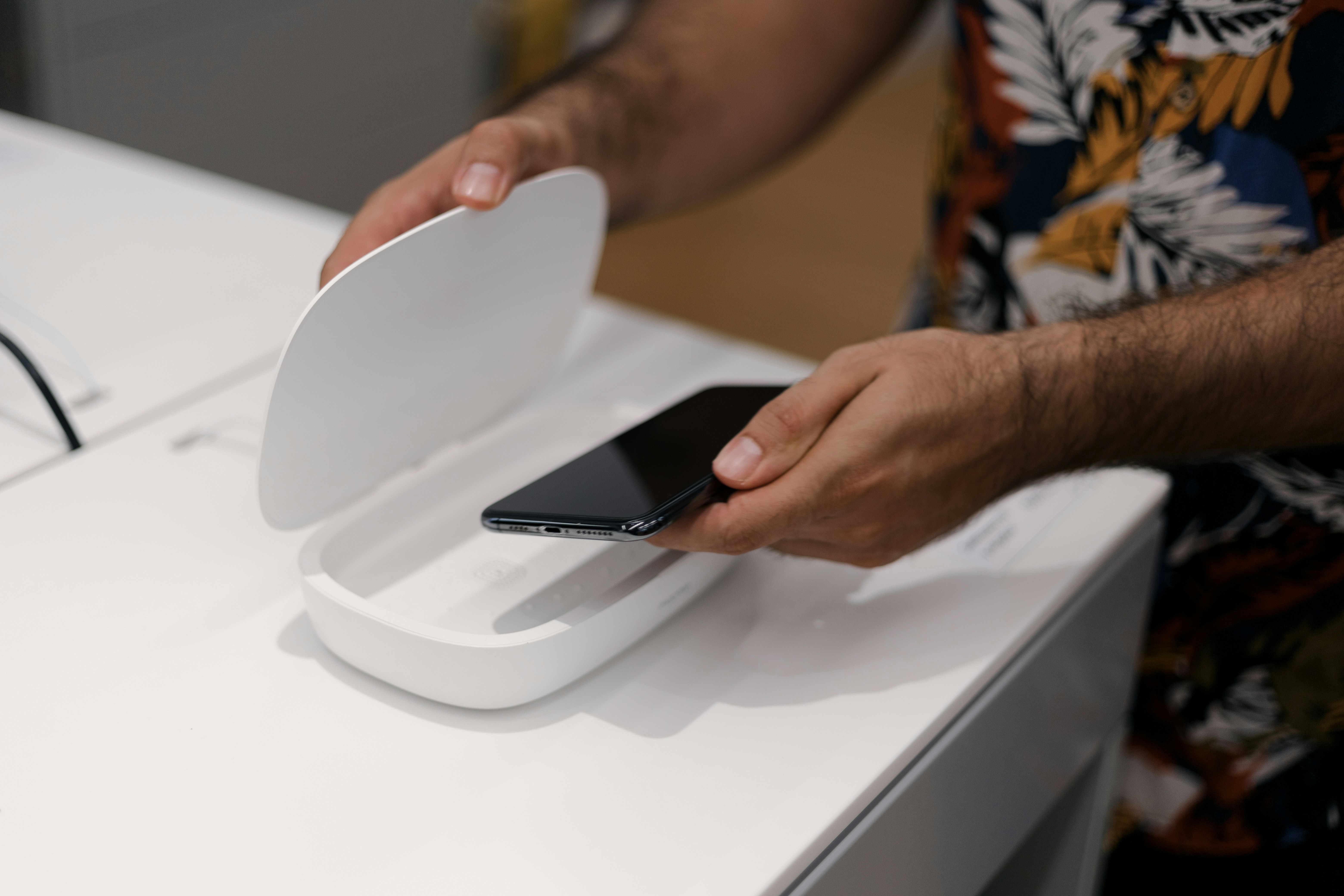 A person placing a smartphone into a sleek, white sanitizing device on a modern surface.