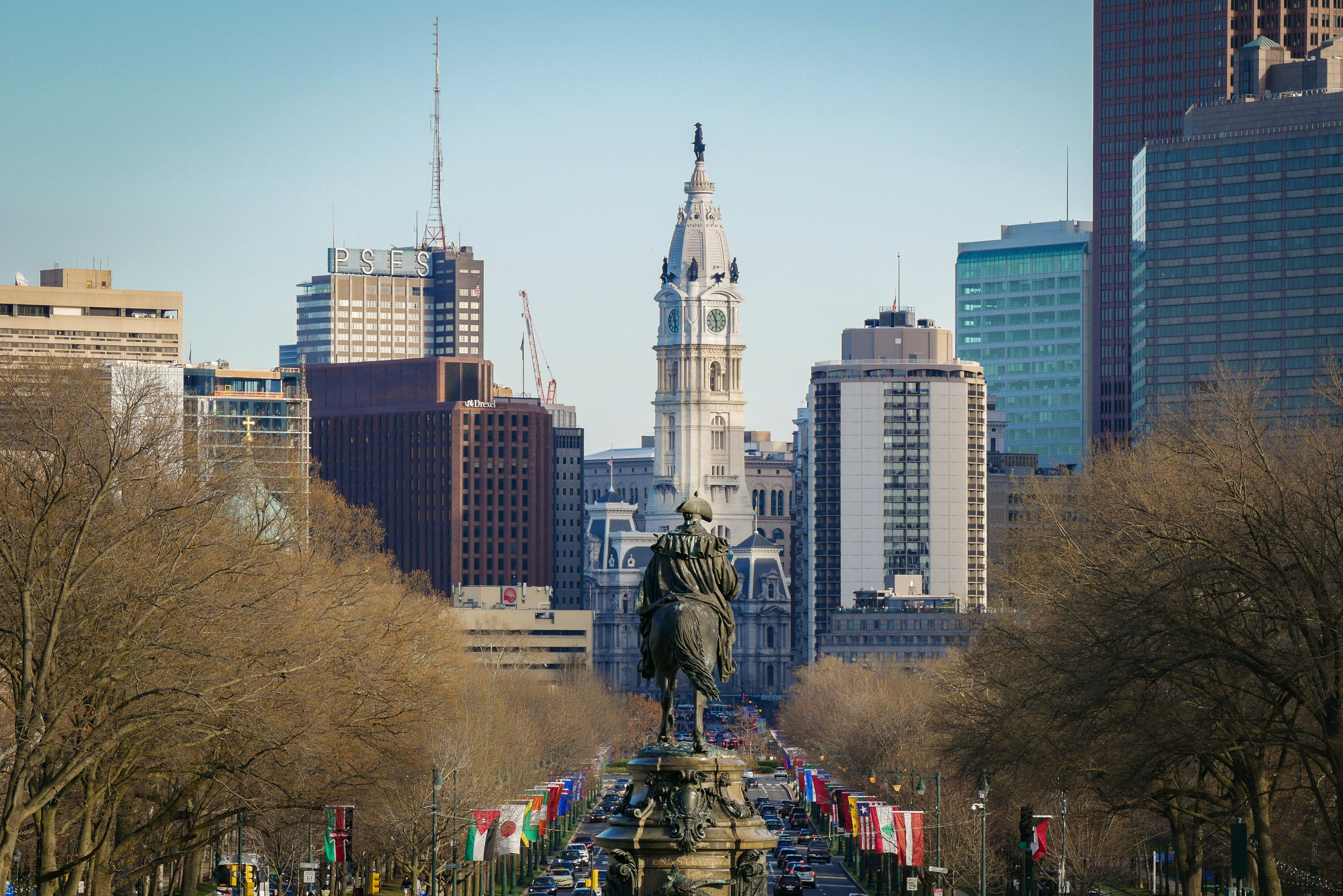 Photograph of an equestrian statue at the head of a tree-lined avenue, with Philadelphia City Hall looming in the distance. Flags line the plaza, adding color to the stately scene.