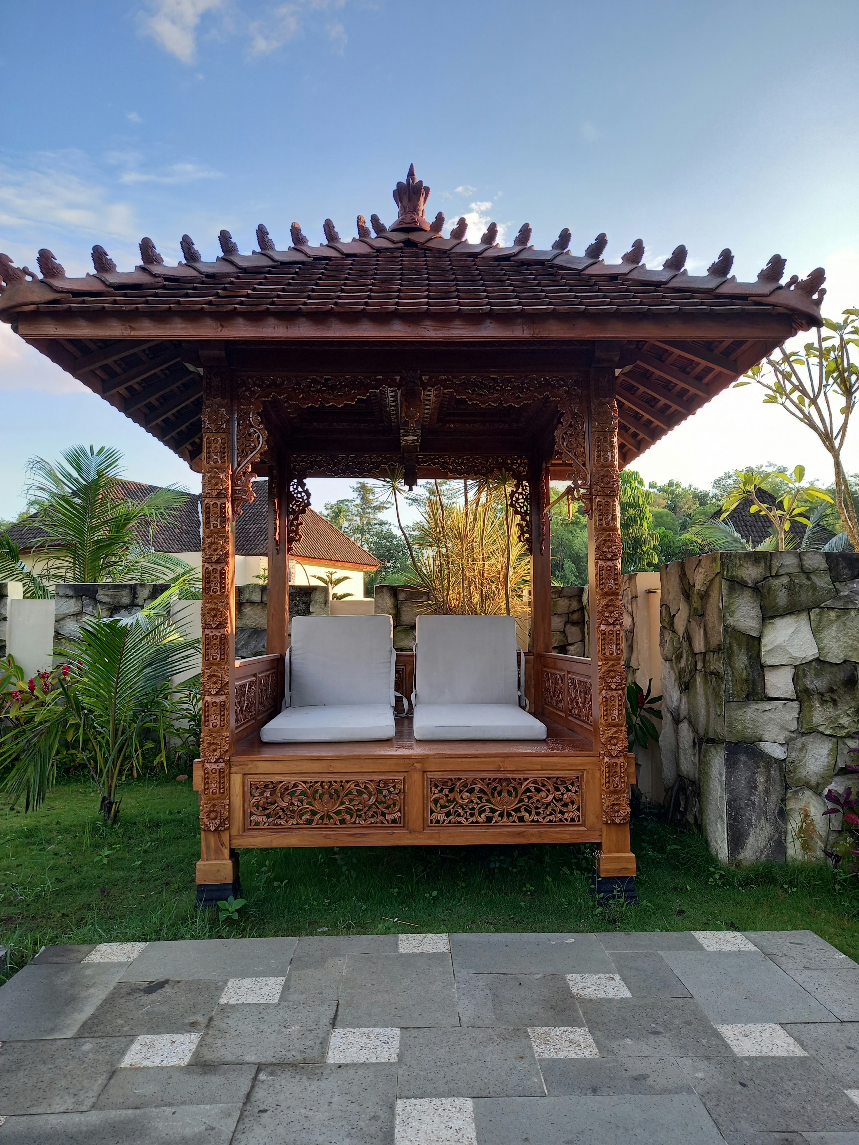 Ornate wooden gazebo with white cushions sits on a stone-paved foreground in a lush garden, framed by stone walls and tropical plants.
