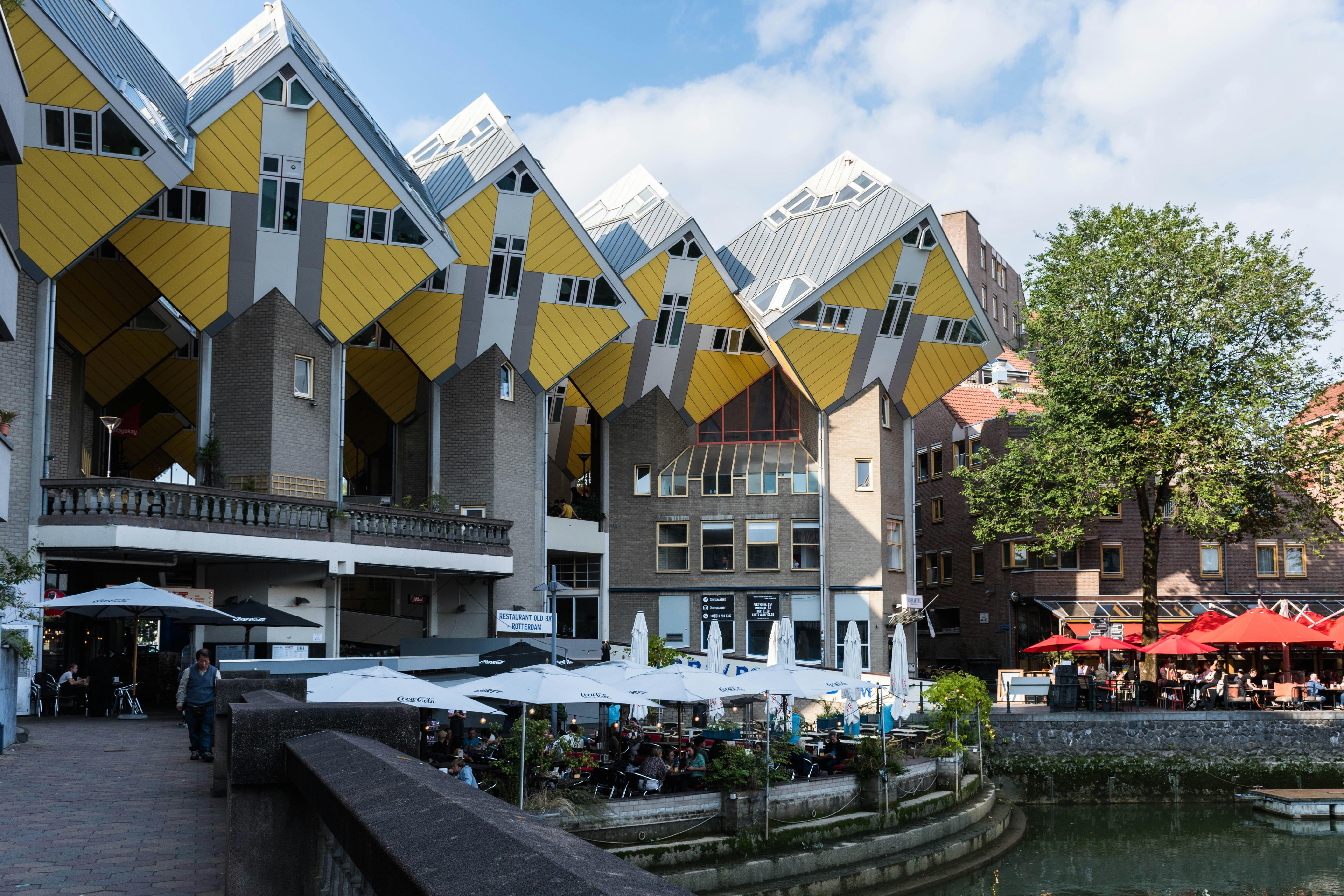 Unique cube-shaped architecture with vibrant yellow accents, surrounded by outdoor dining spaces and lush greenery along a waterway.