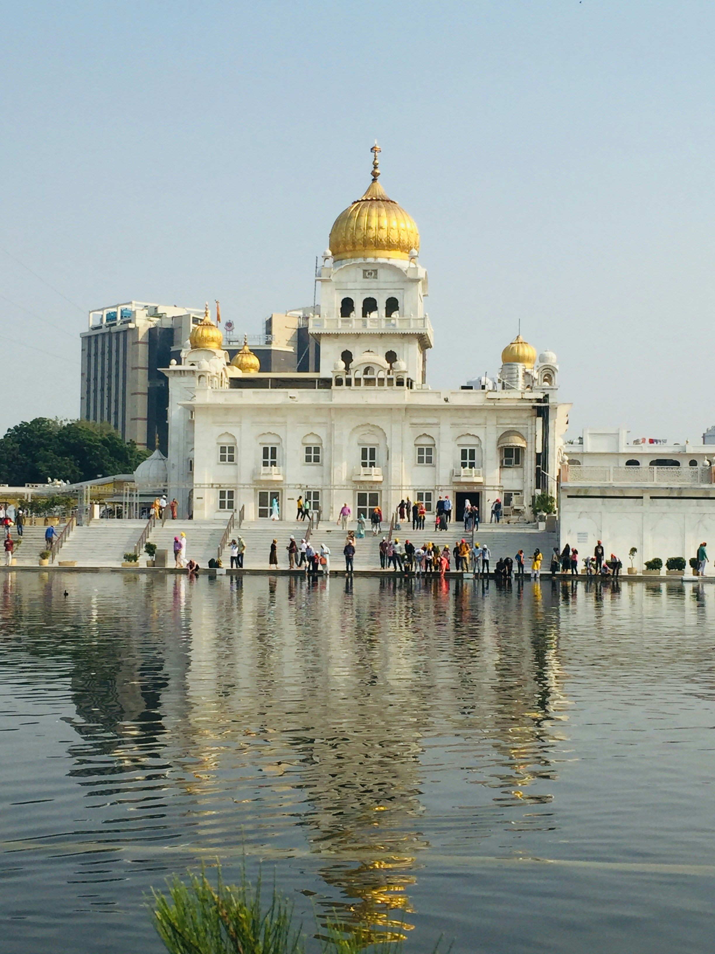 A majestic white temple with golden domes stands beside a tranquil lake, surrounded by visitors enjoying the serene atmosphere.