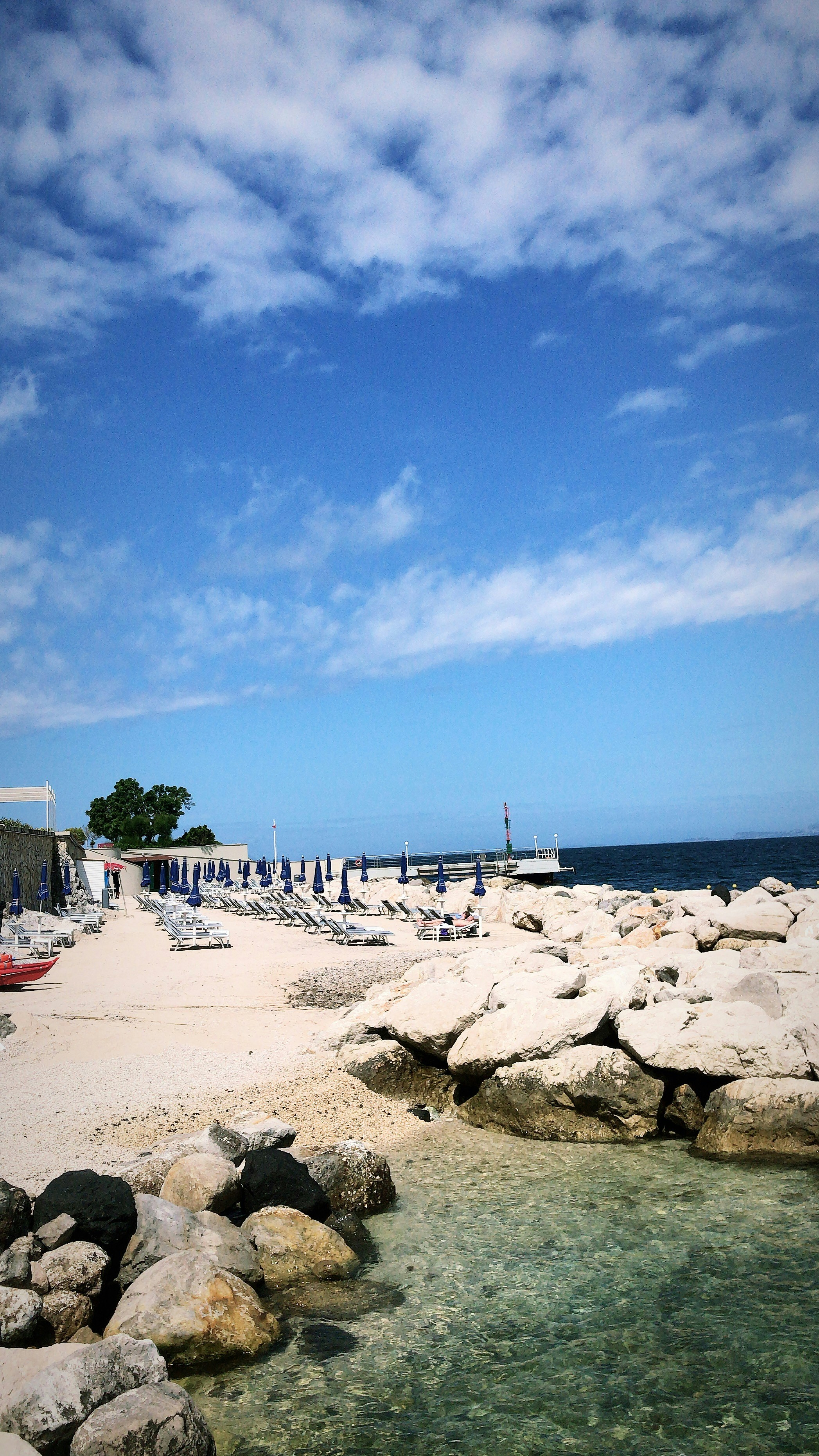 A tranquil beach scene featuring lounge chairs and umbrellas along the shoreline, with clear water and rocky outcrops in the foreground.