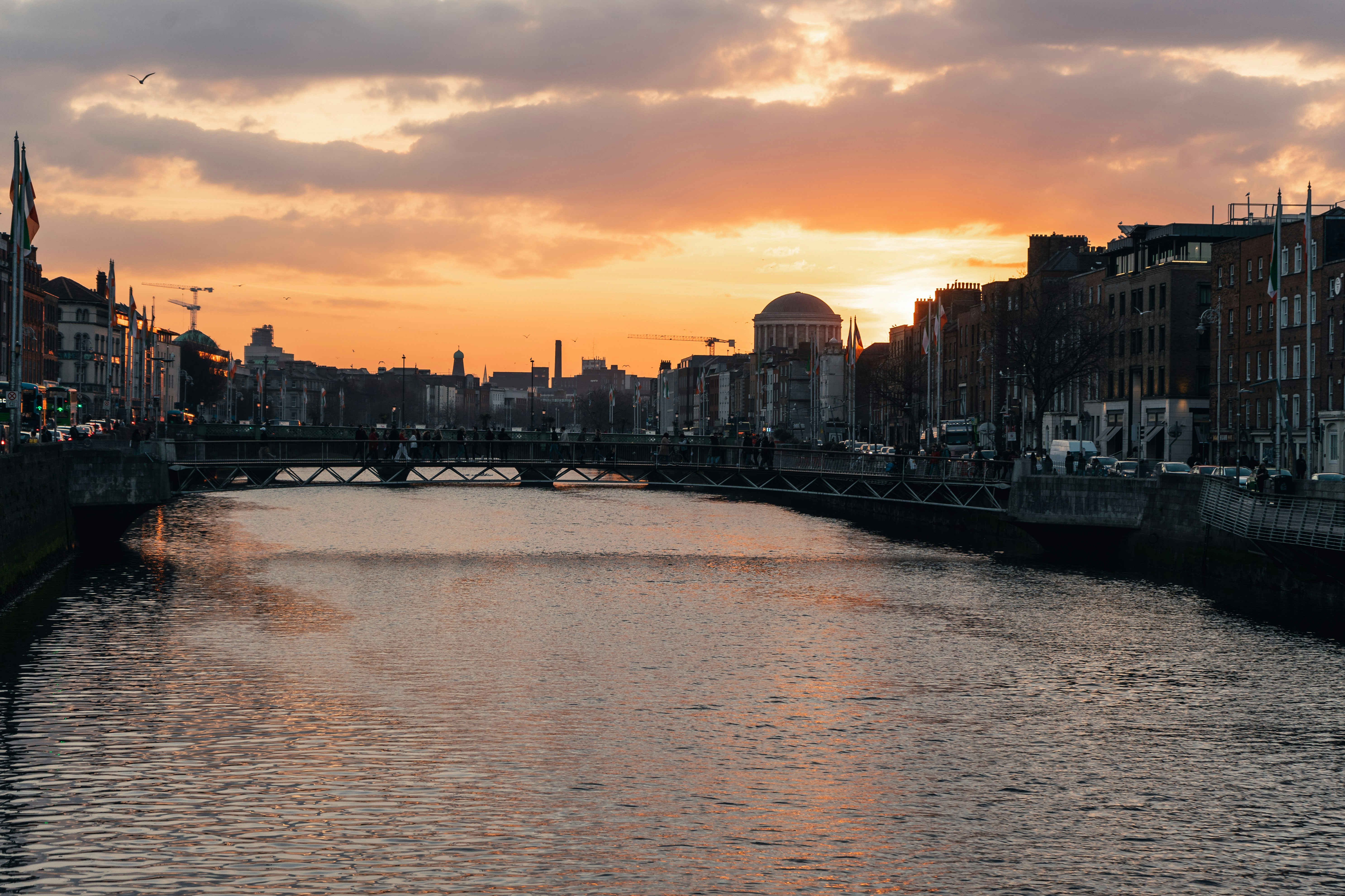 The River Liffey glimmers under a vibrant sunset, framed by historic buildings and a bustling bridge. The scene captures the essence of Dublin's evening charm.
