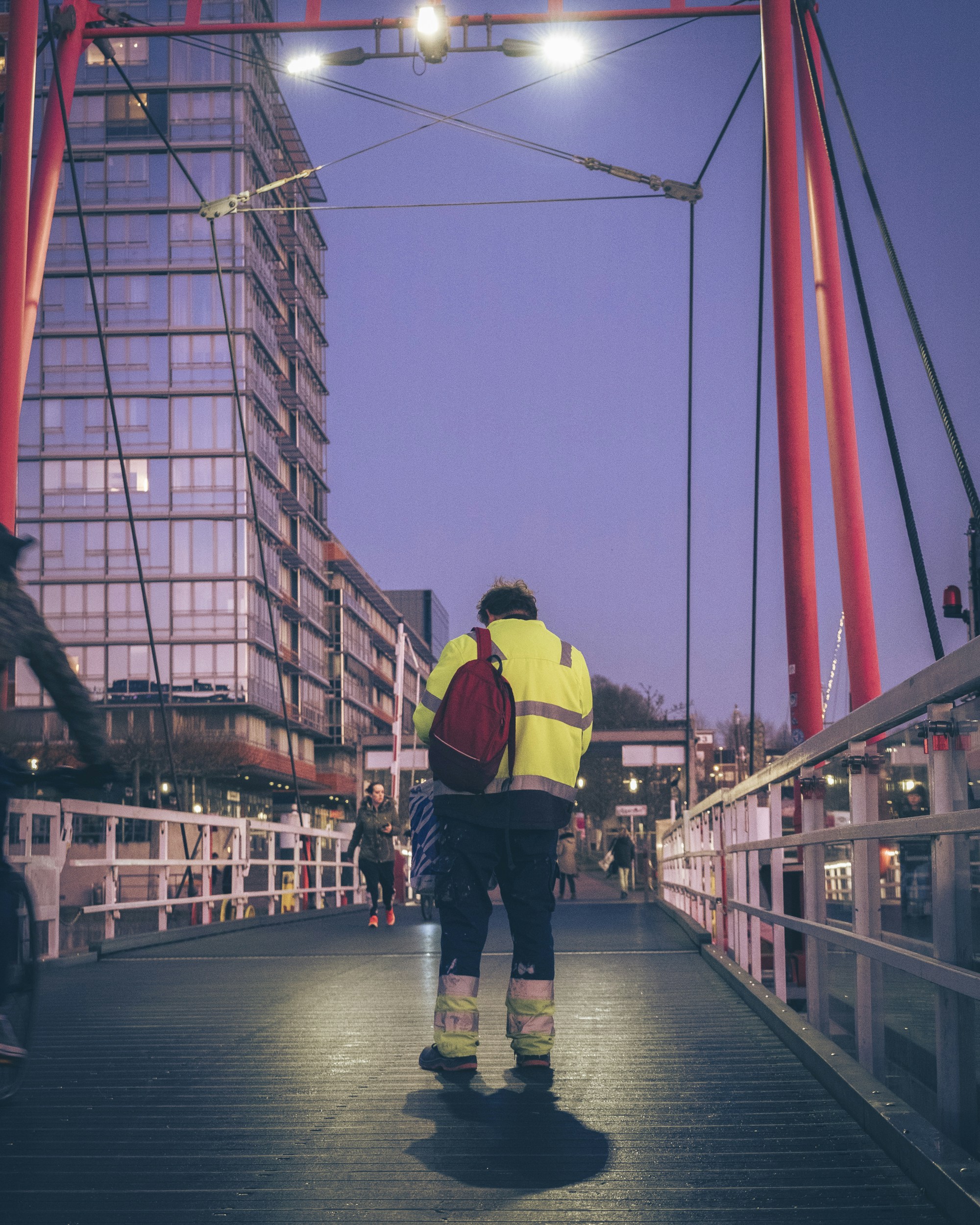 A worker in a reflective jacket stands on a bridge, illuminated by overhead lights, as pedestrians pass by in the fading twilight.