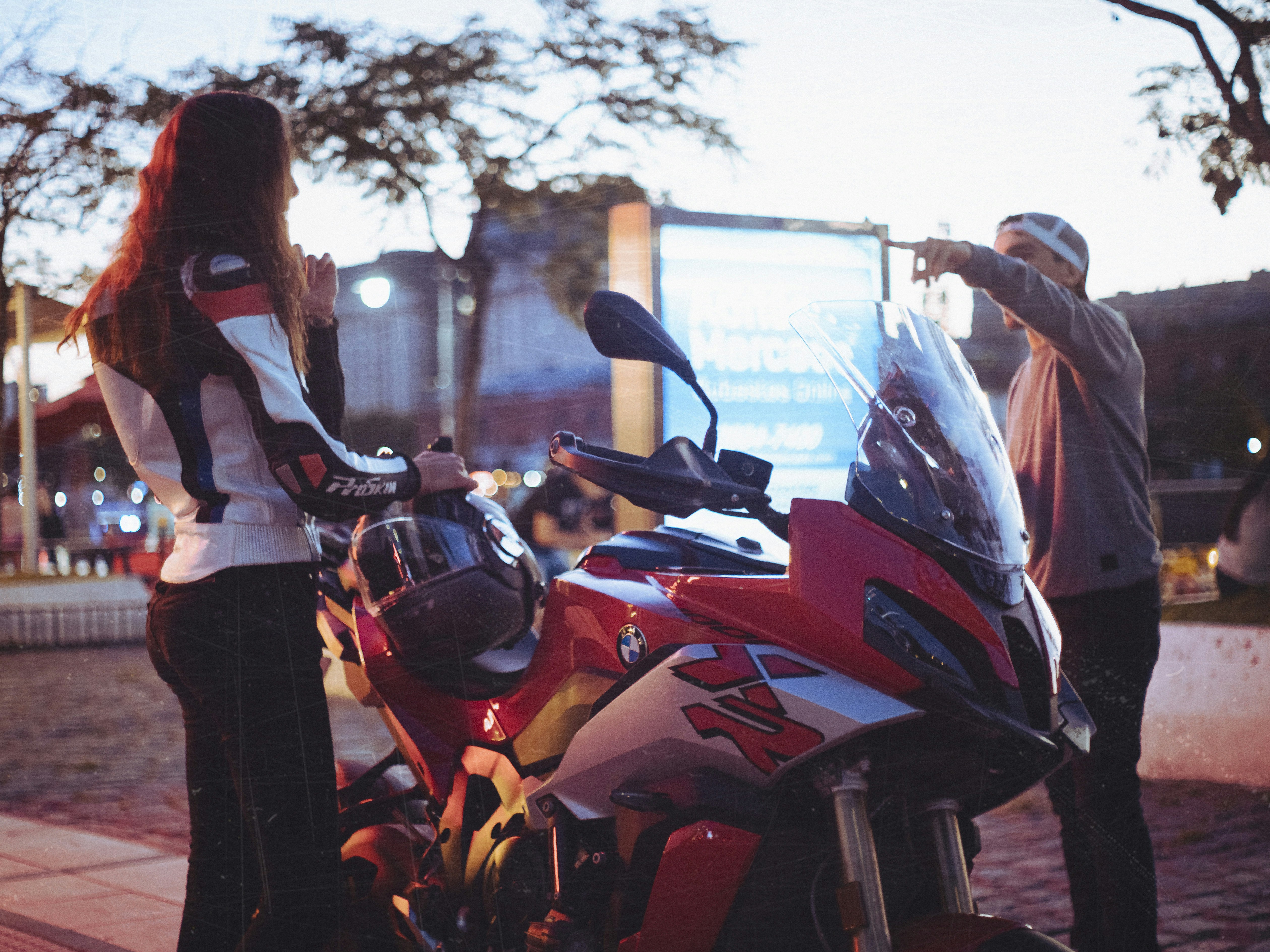 A motorcyclist in gear stands beside a vibrant motorcycle, engaging in conversation with a companion as dusk settles in. The scene captures the camaraderie and passion for biking.
